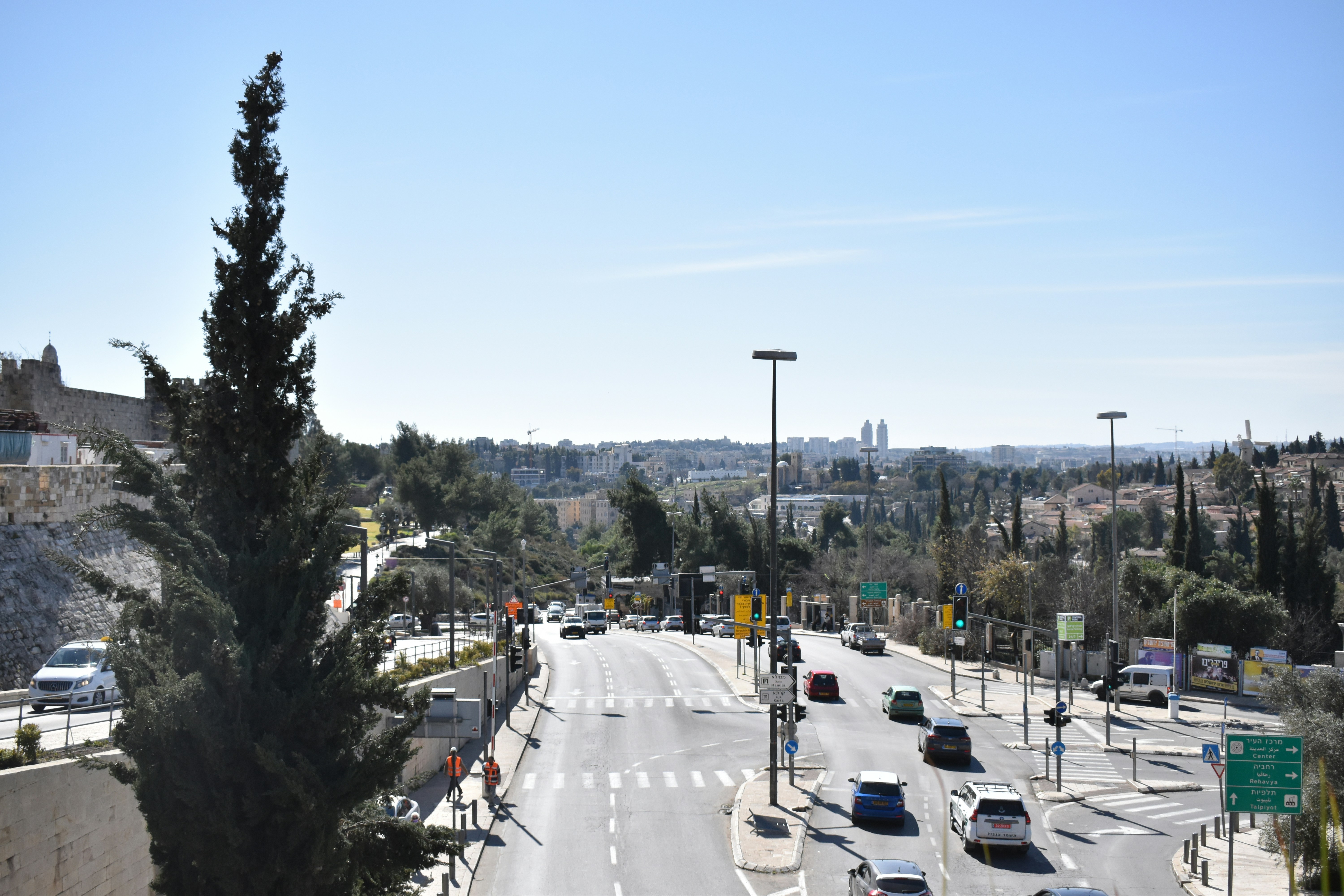 A busy intersection in Jerusalem showcasing traffic, trees, and distant hills under a clear blue sky.