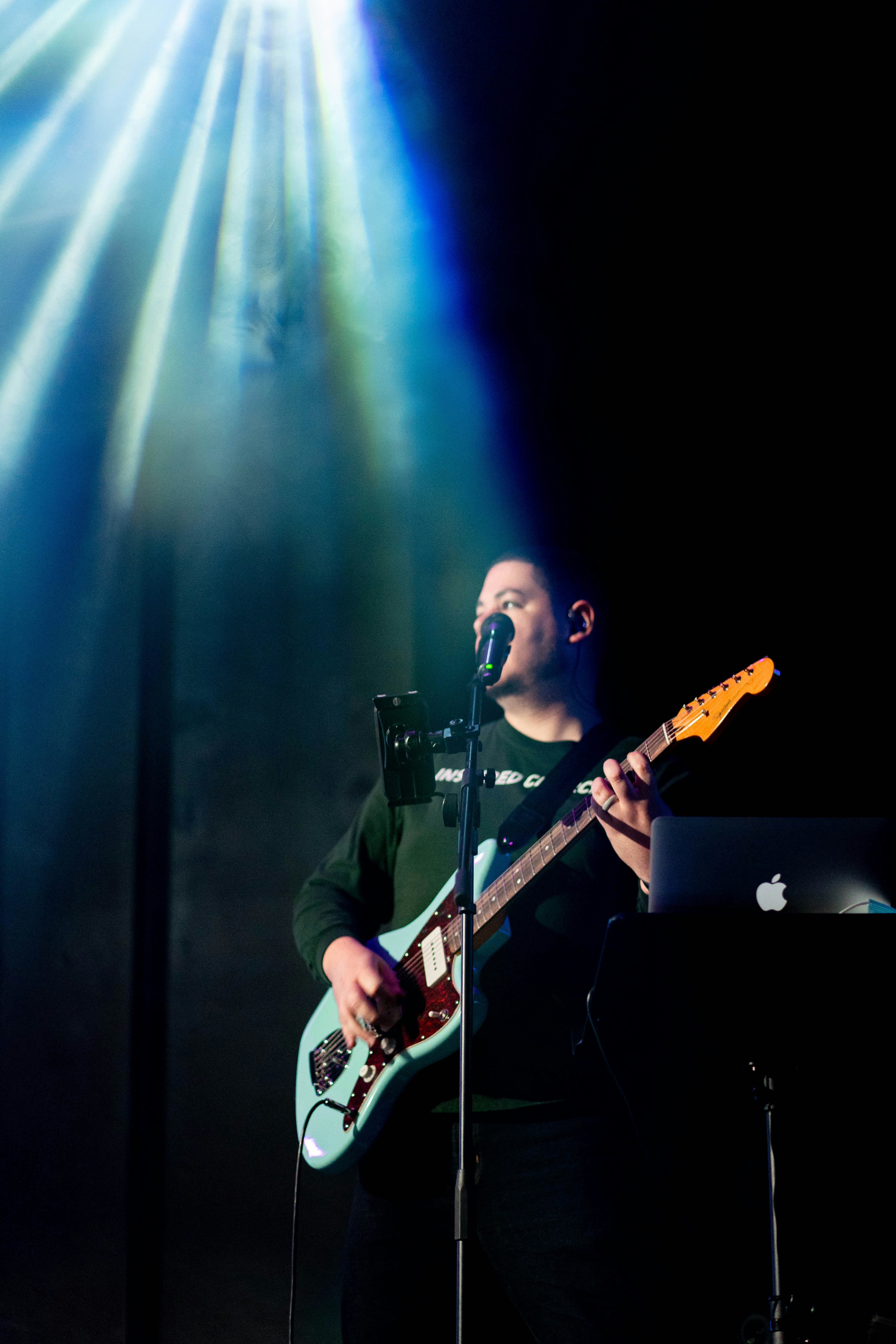 Musician passionately playing an electric guitar under dramatic stage lighting, with a laptop visible nearby.