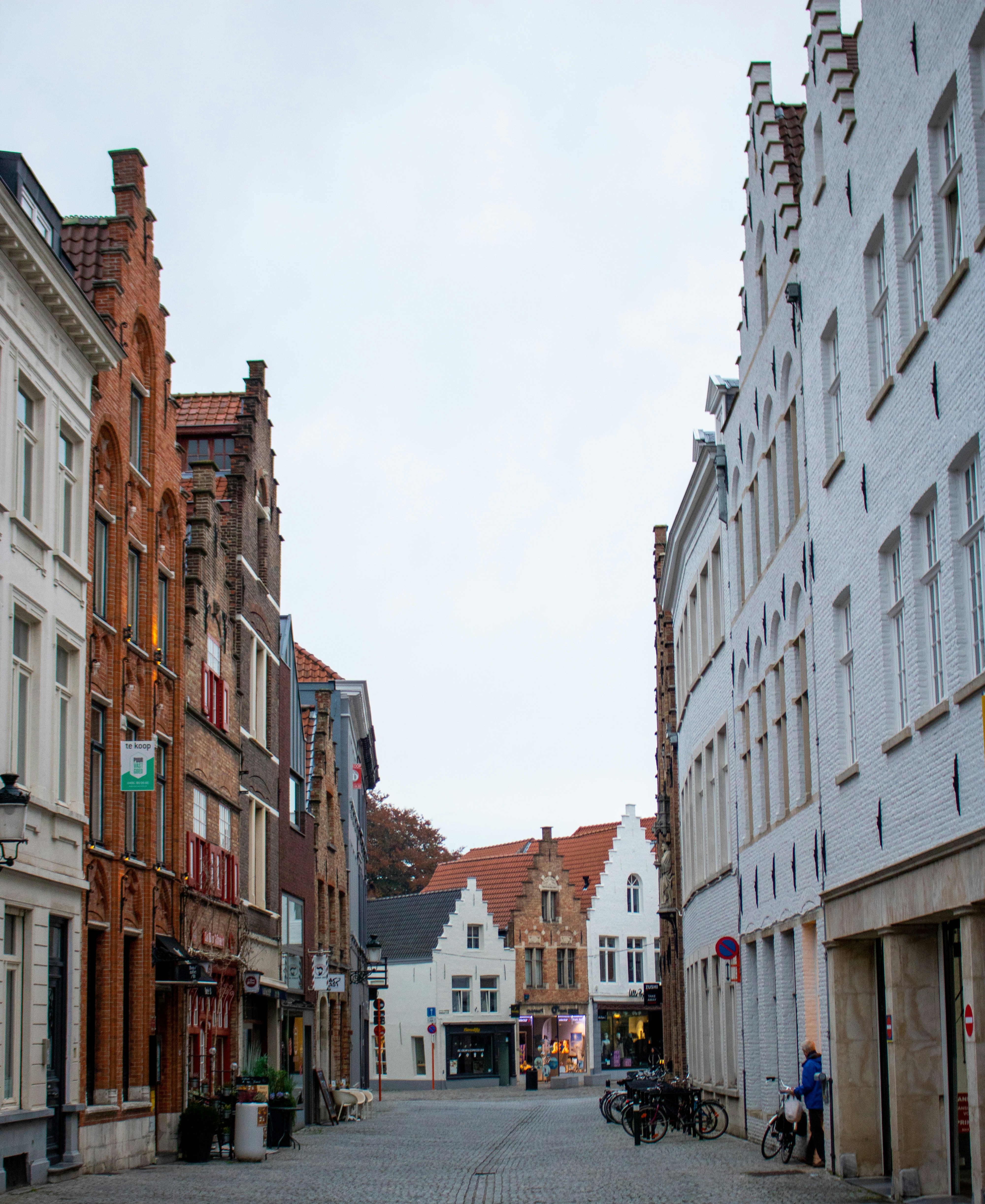 Charming street lined with historic buildings showcasing a blend of architectural styles, leading to a quaint square. A cyclist pauses, adding life to the serene scene.