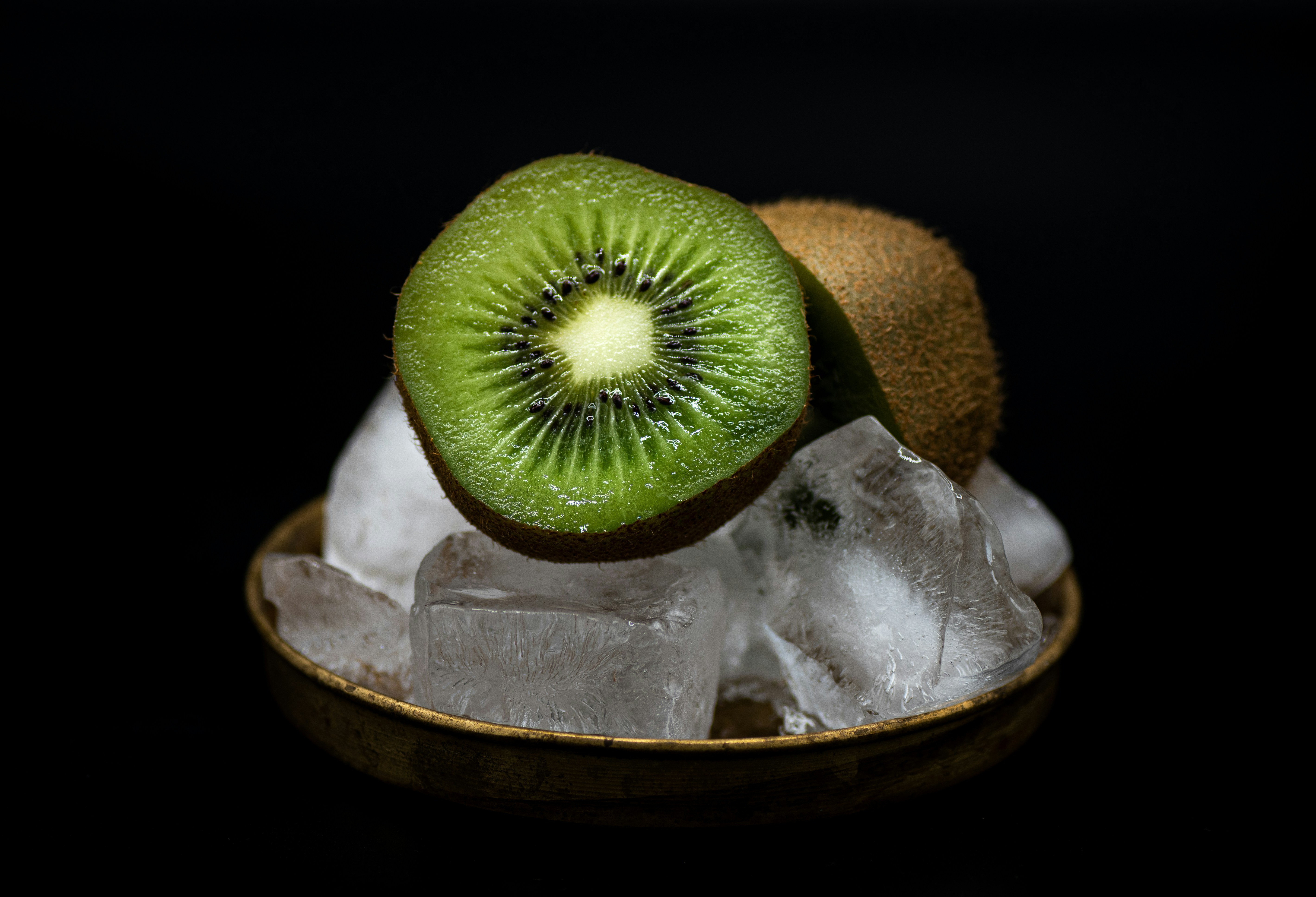 A halved kiwi rests atop a bed of ice cubes in a shallow dish, showcasing its vibrant green flesh and unique texture against a dark background.