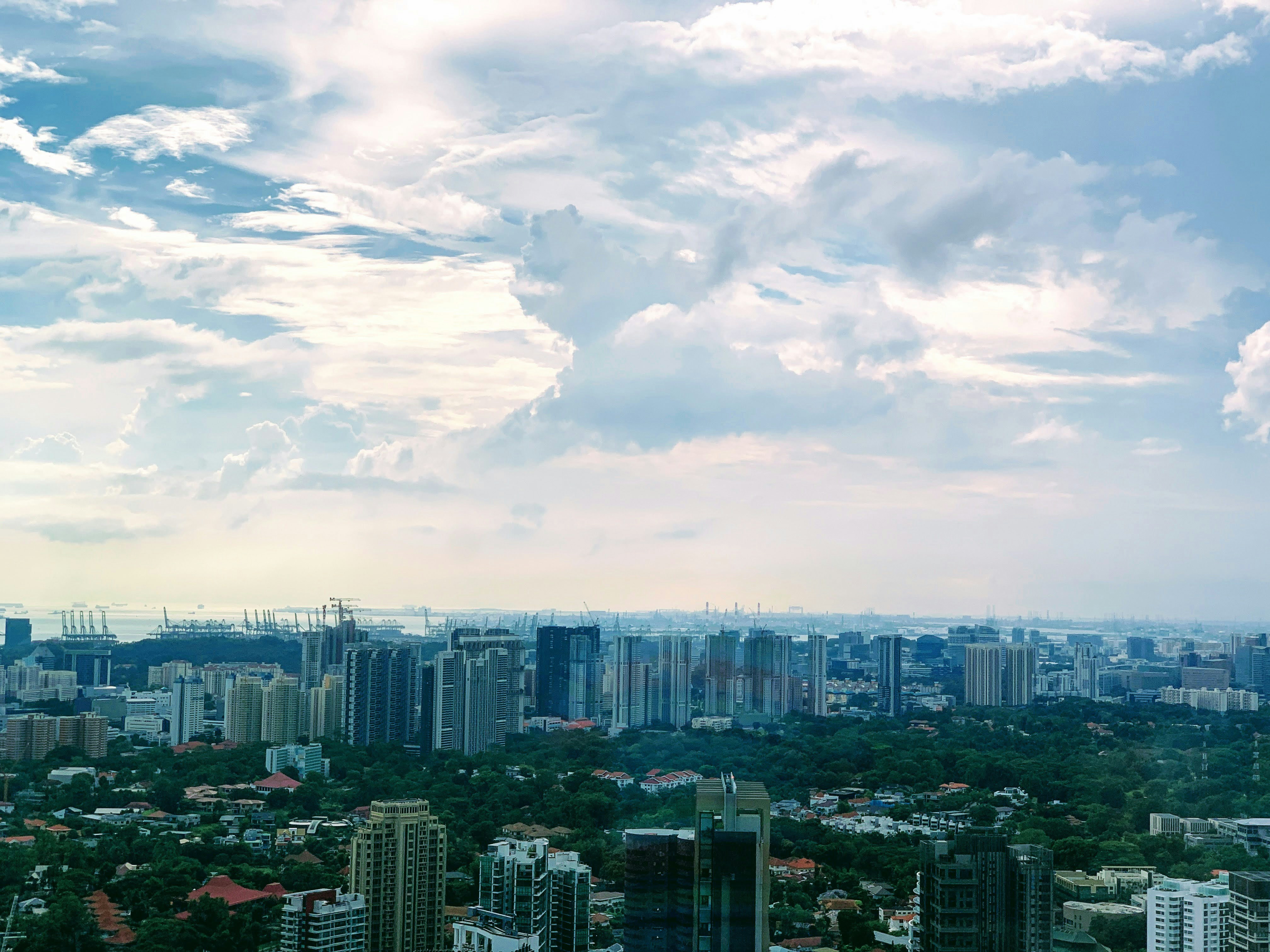 City skyline from a high vantage point with layered clouds and distant harbor cranes against a bright sky.