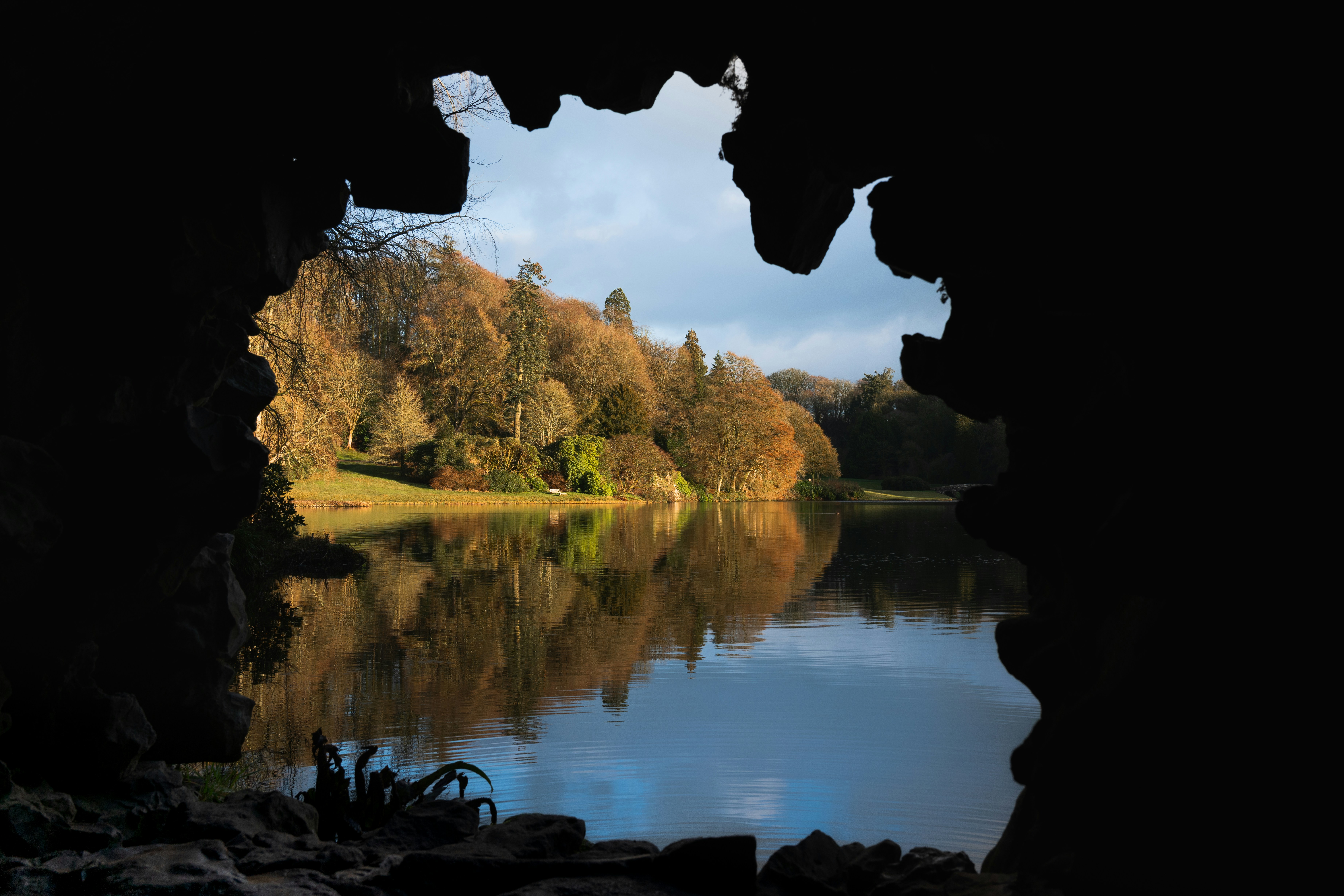 Scenic view of a serene lake surrounded by autumn foliage, framed by the jagged edges of a stone structure. The calm waters mirror the vibrant colors of the landscape.