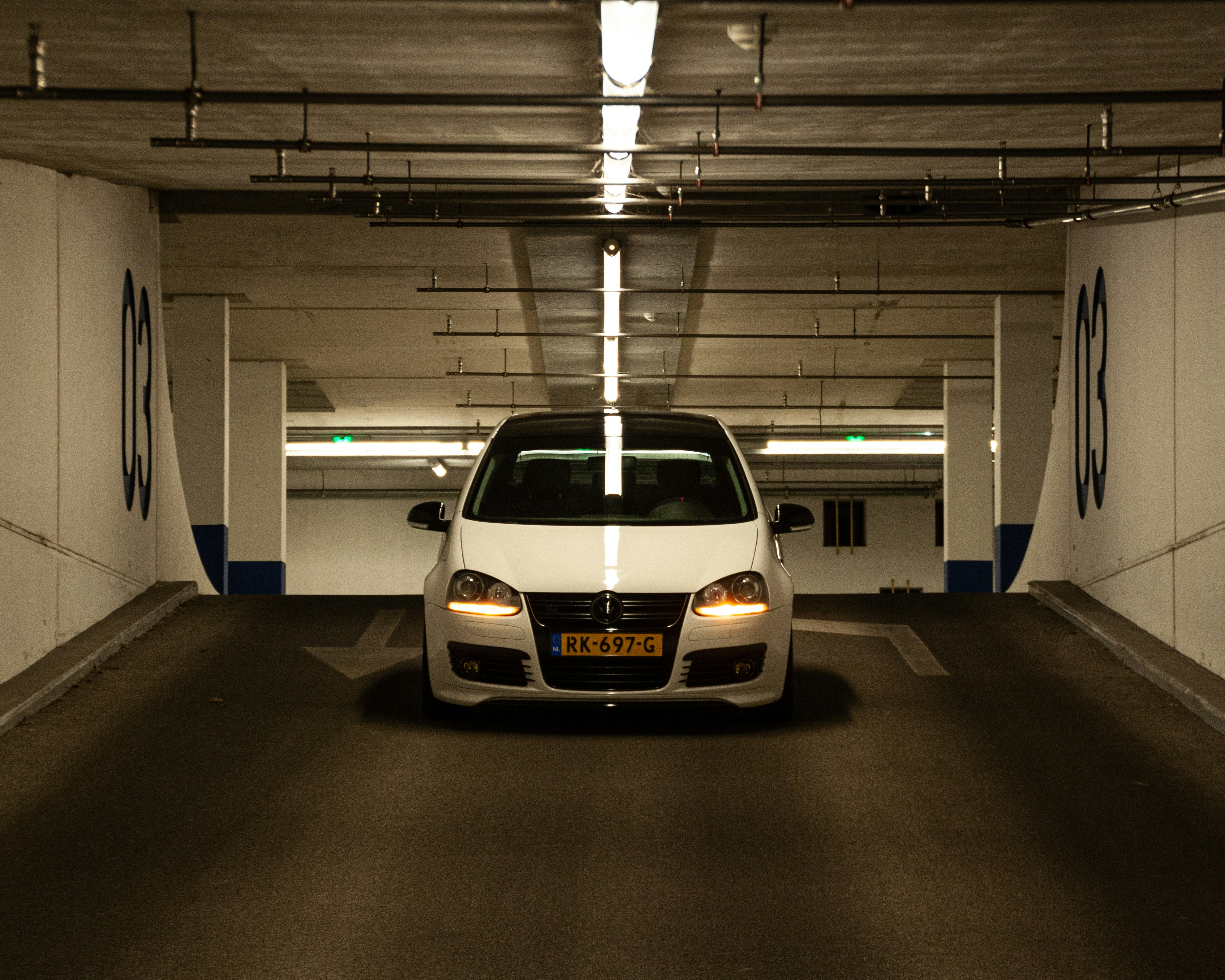 A white car parked in an underground parking garage, illuminated by overhead lights and flanked by stark white walls marked with numbers.