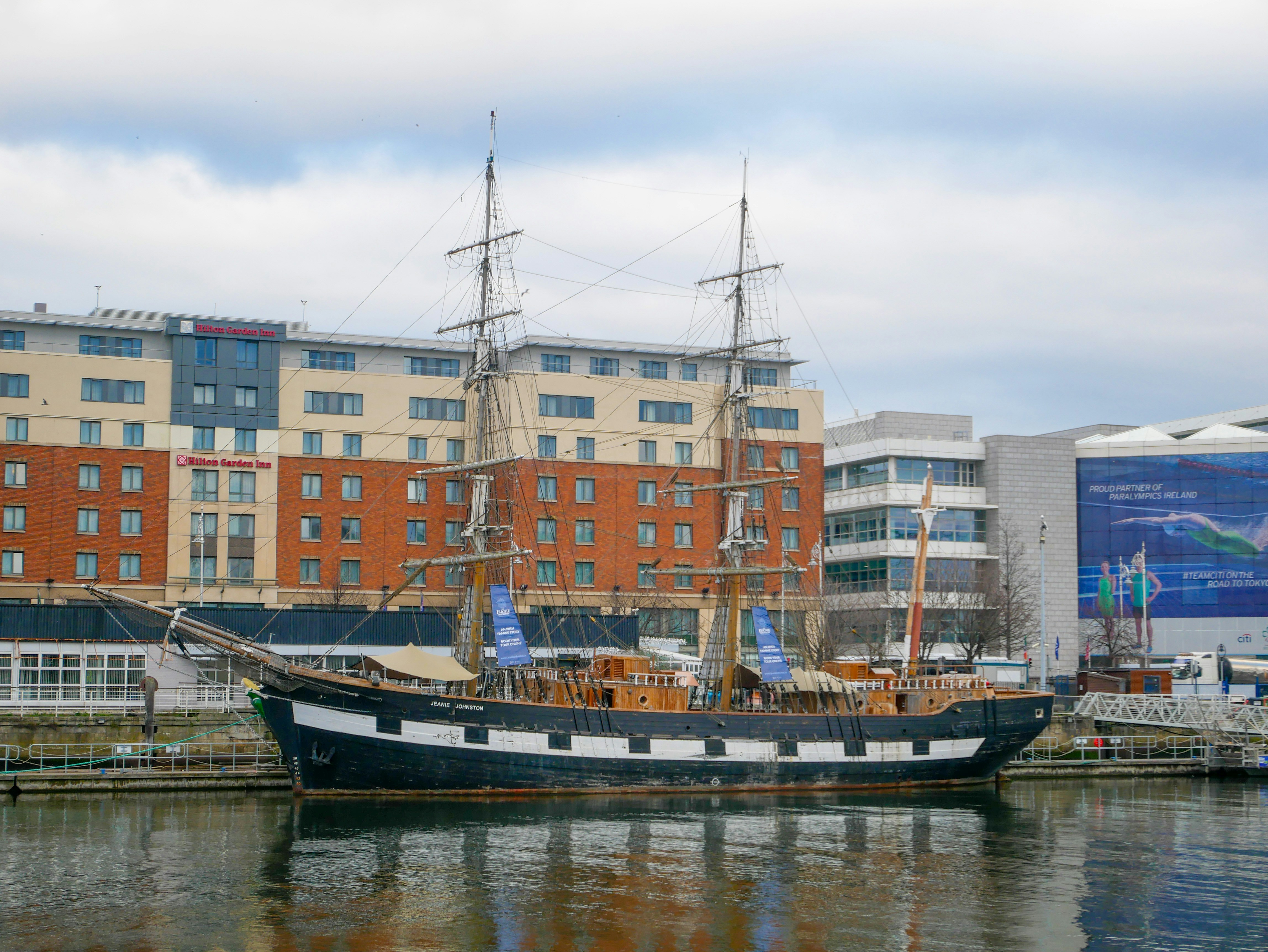 Historic ship docked alongside modern buildings, reflecting the blend of tradition and contemporary architecture in a bustling waterfront.