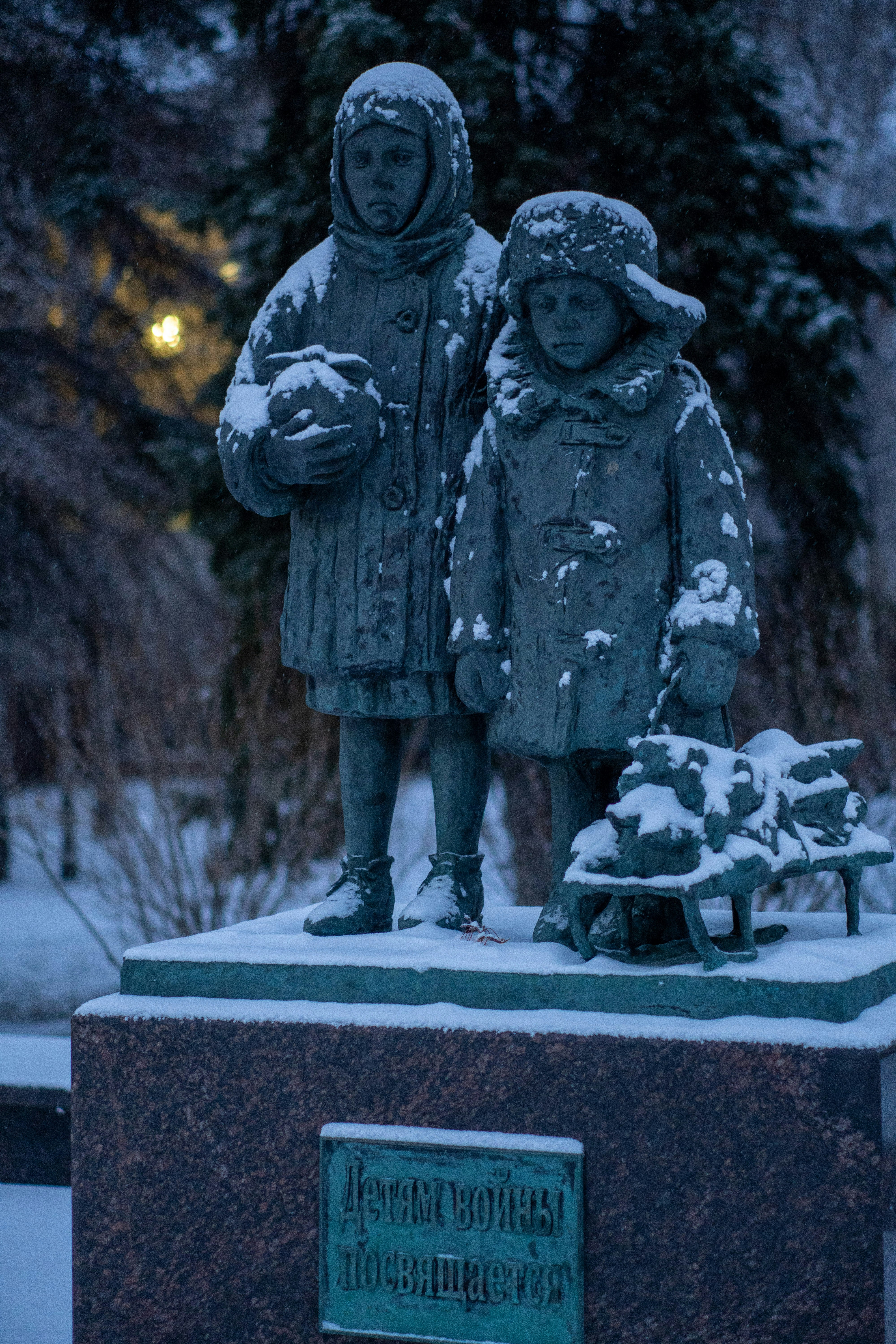 Bronze statue of two children in winter attire, standing together with snow covering them, commemorating the impact of war on youth. The inscription honors the memory of children affected by conflict.