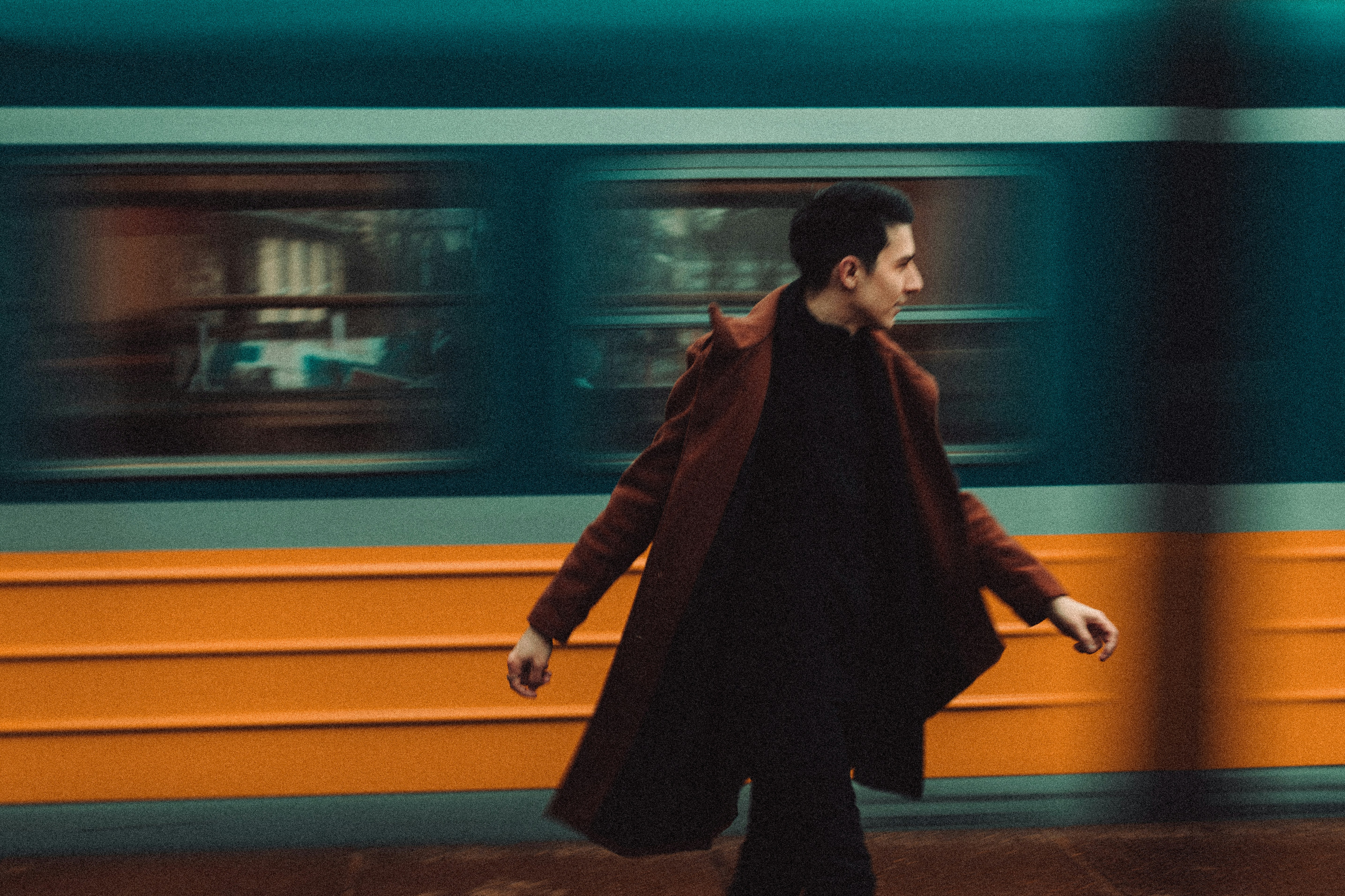 A figure in a long coat strides confidently past a blurred train, capturing the essence of urban life in motion.