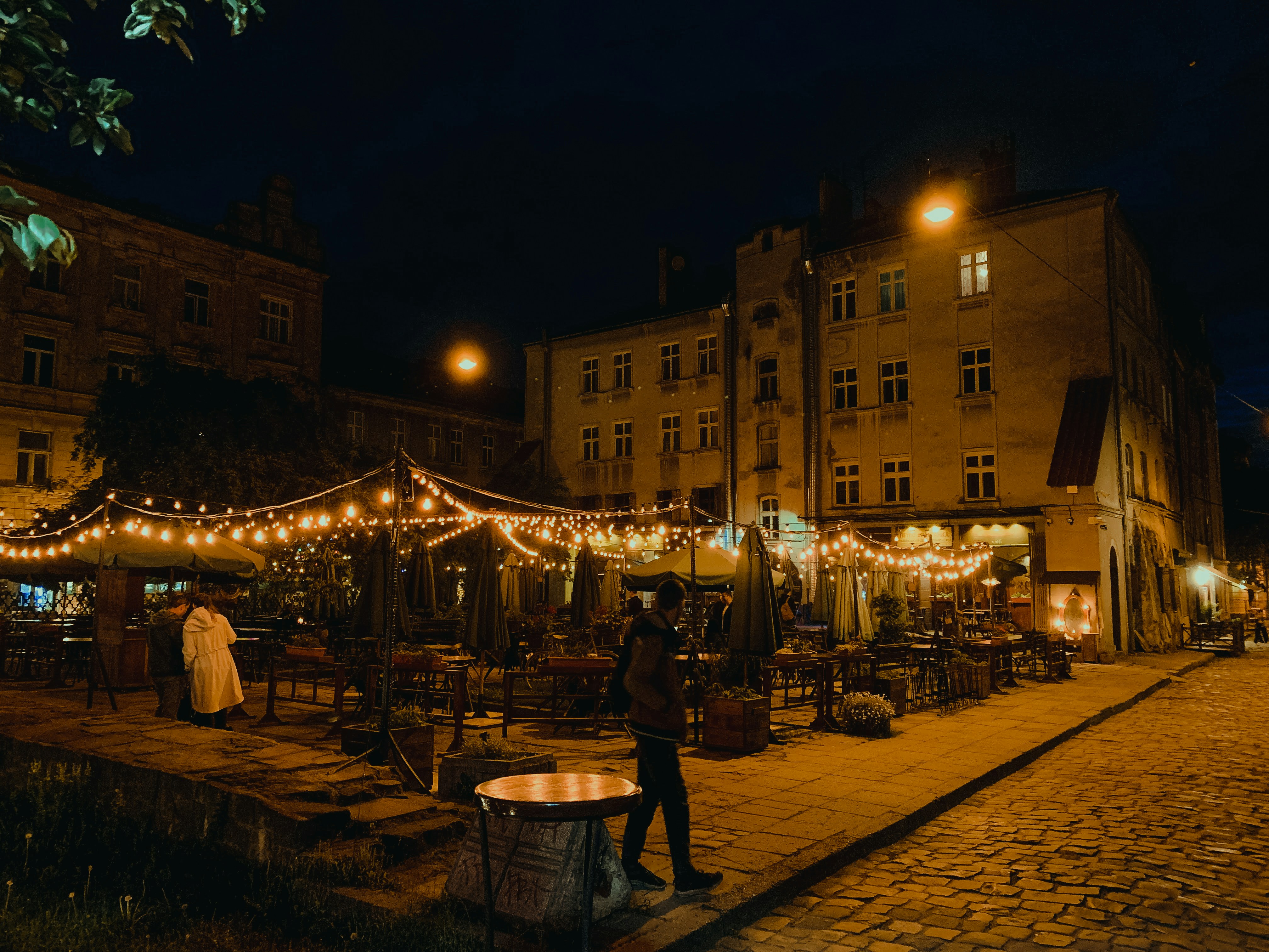 Person walking past a warmly lit outdoor cafe on a cobblestone street at night.
