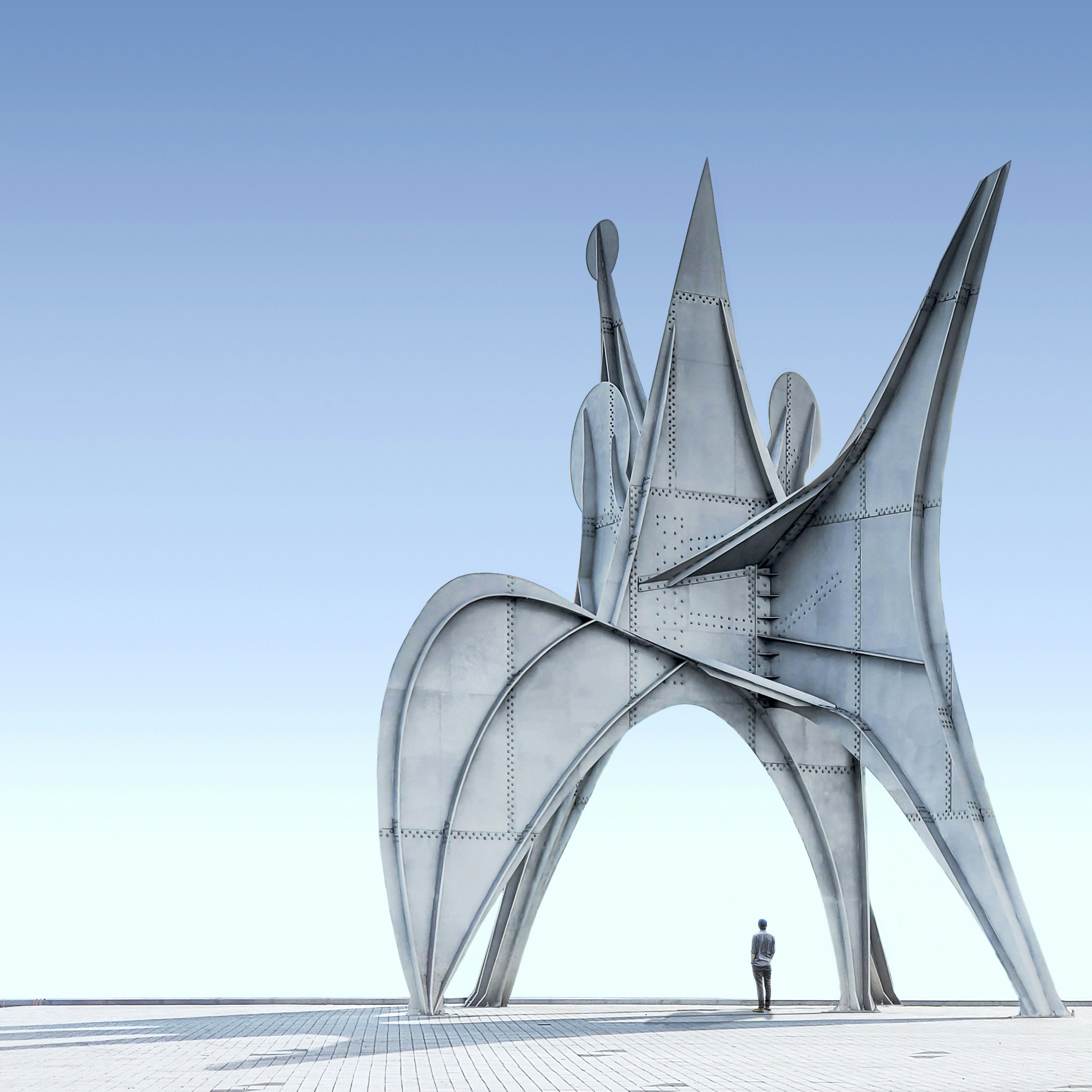 A monumental steel sculpture with sweeping curves rises on a wide plaza beneath a pale blue gradient sky, with a lone figure for scale.