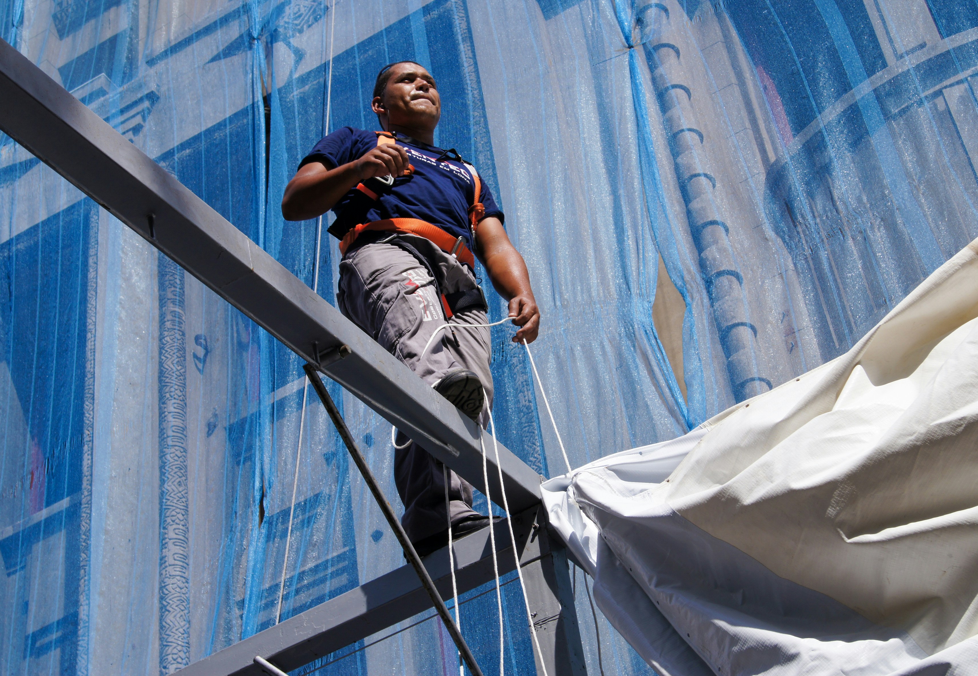 Worker in harness atop a high structure surrounded by blue construction netting.
