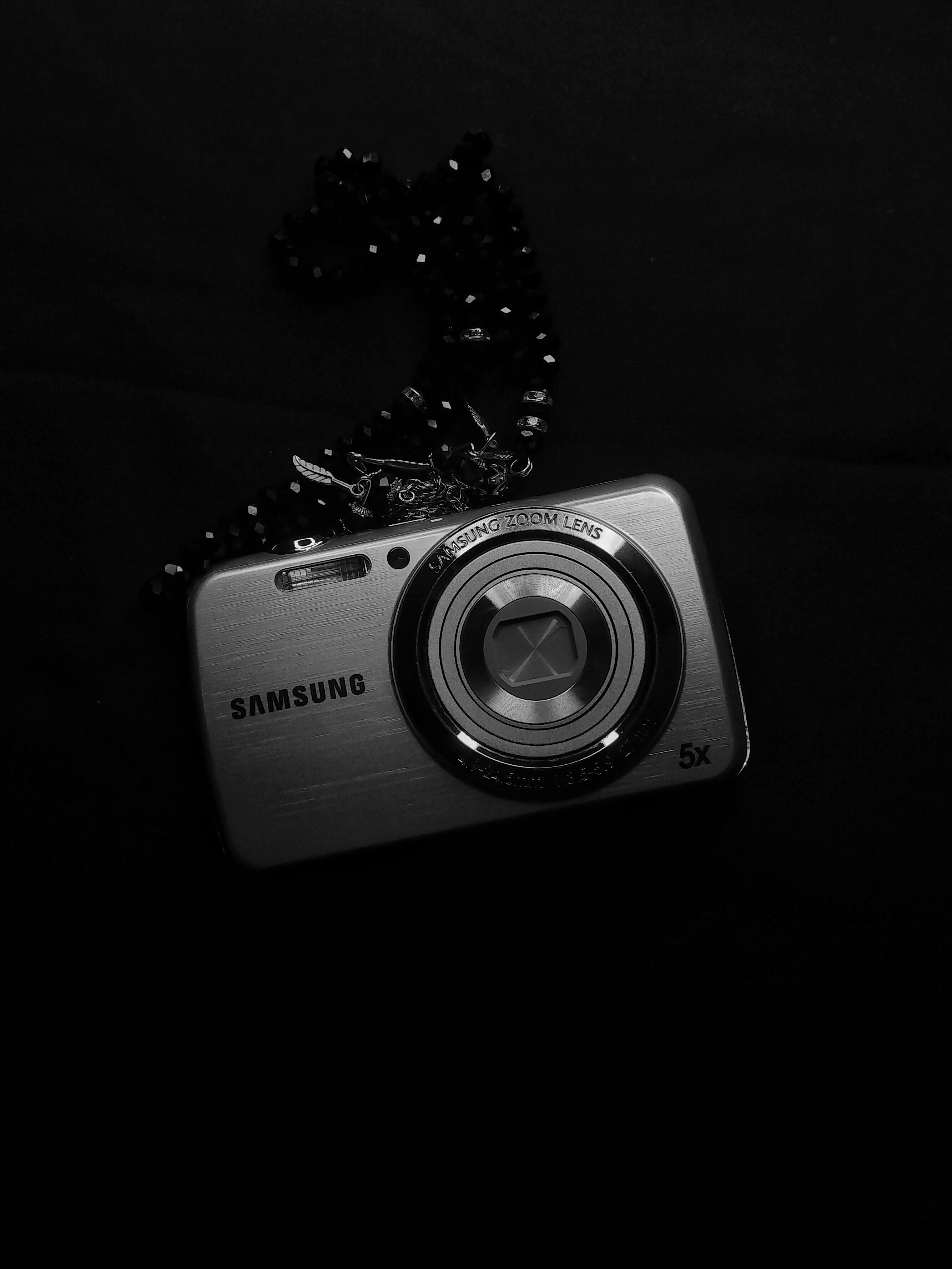 Compact silver camera with a zoom lens and decorative chain, resting on a black surface.