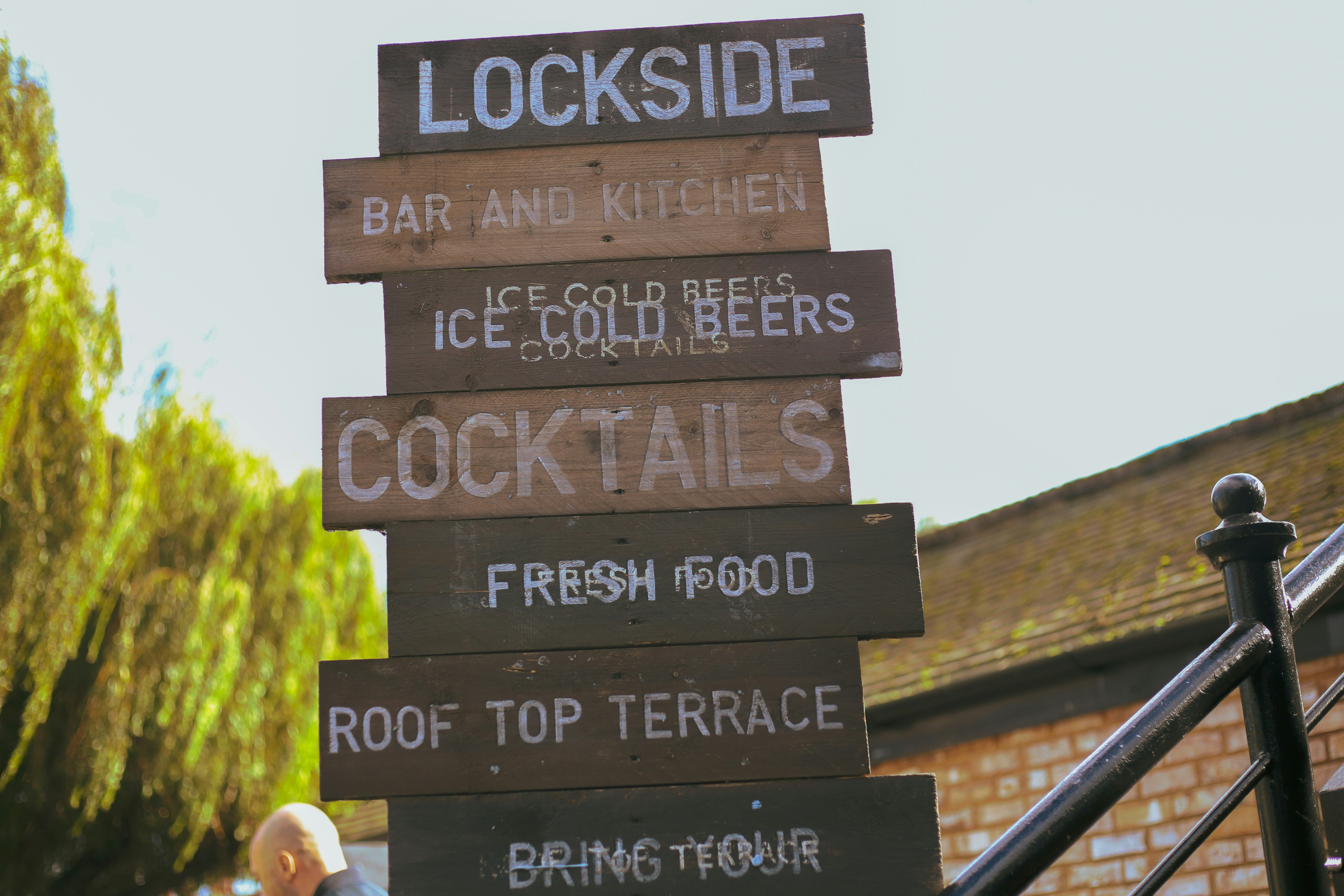 Wooden signpost displaying various offerings of a bar and kitchen, including cocktails and fresh food.