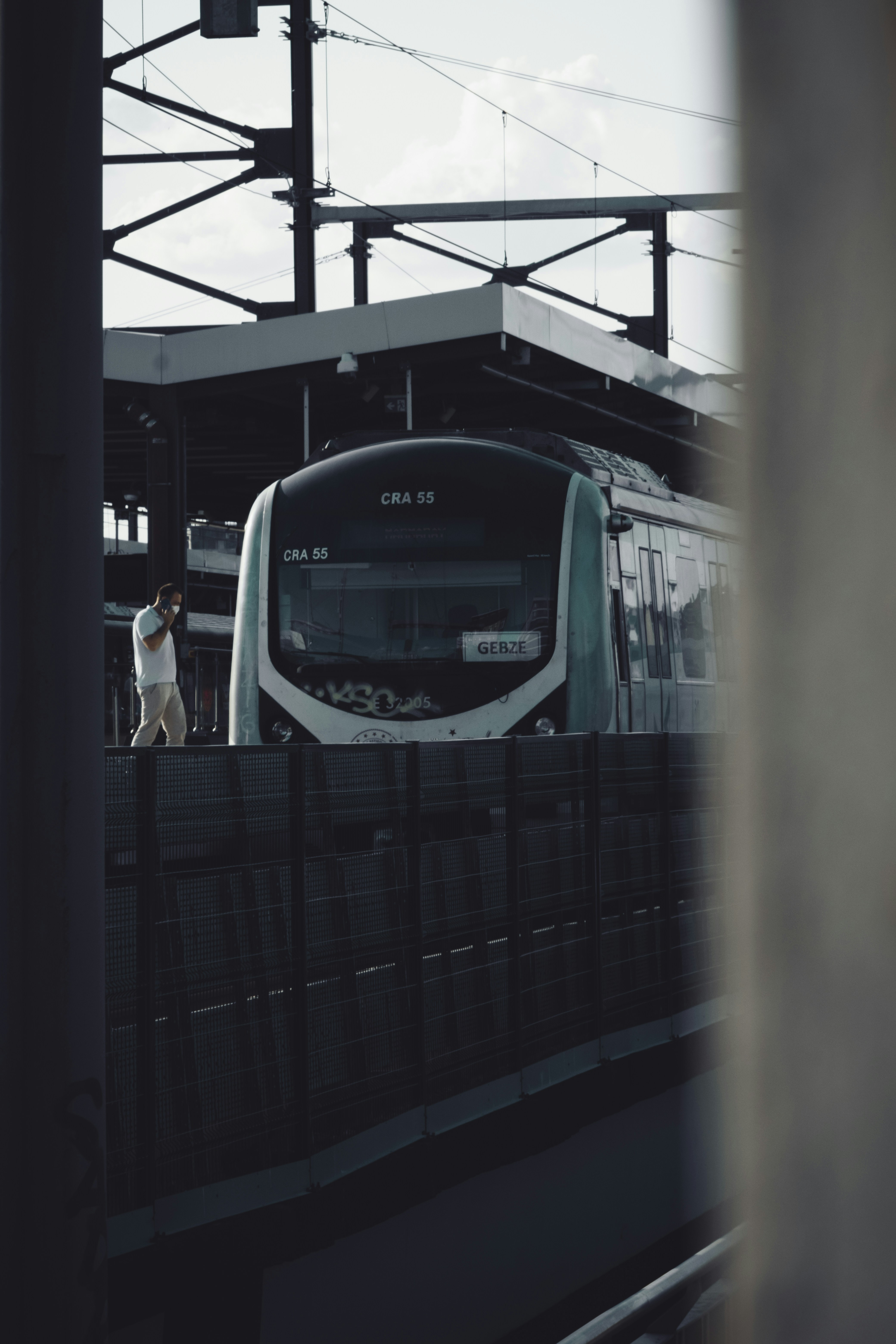 A train labeled CRA 55 approaches the station as a passenger walks along the platform, framed by industrial elements.