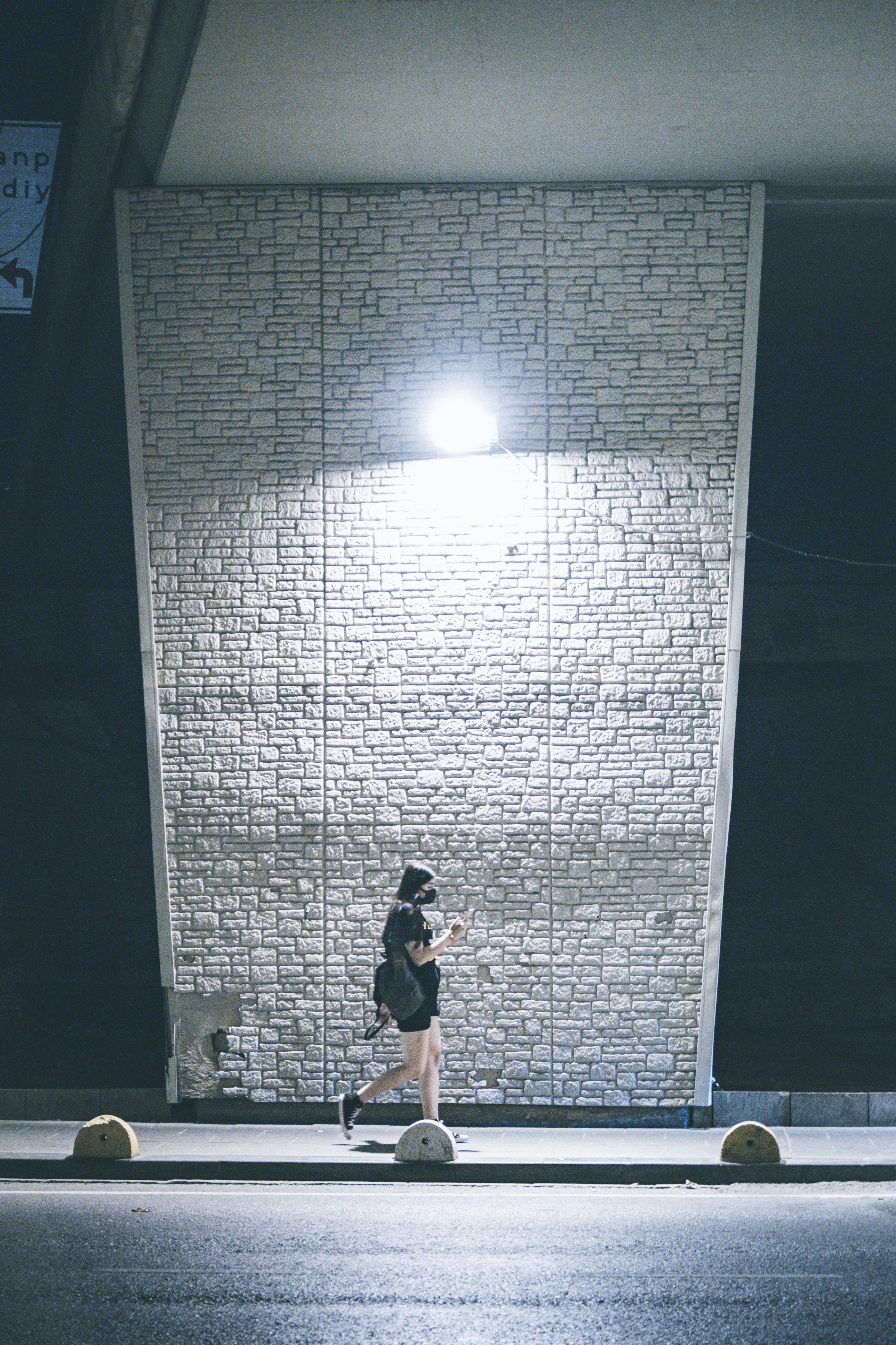 A solitary figure walks along a dimly lit street, illuminated by a bright overhead light against a textured wall. The scene captures the essence of urban life at night.