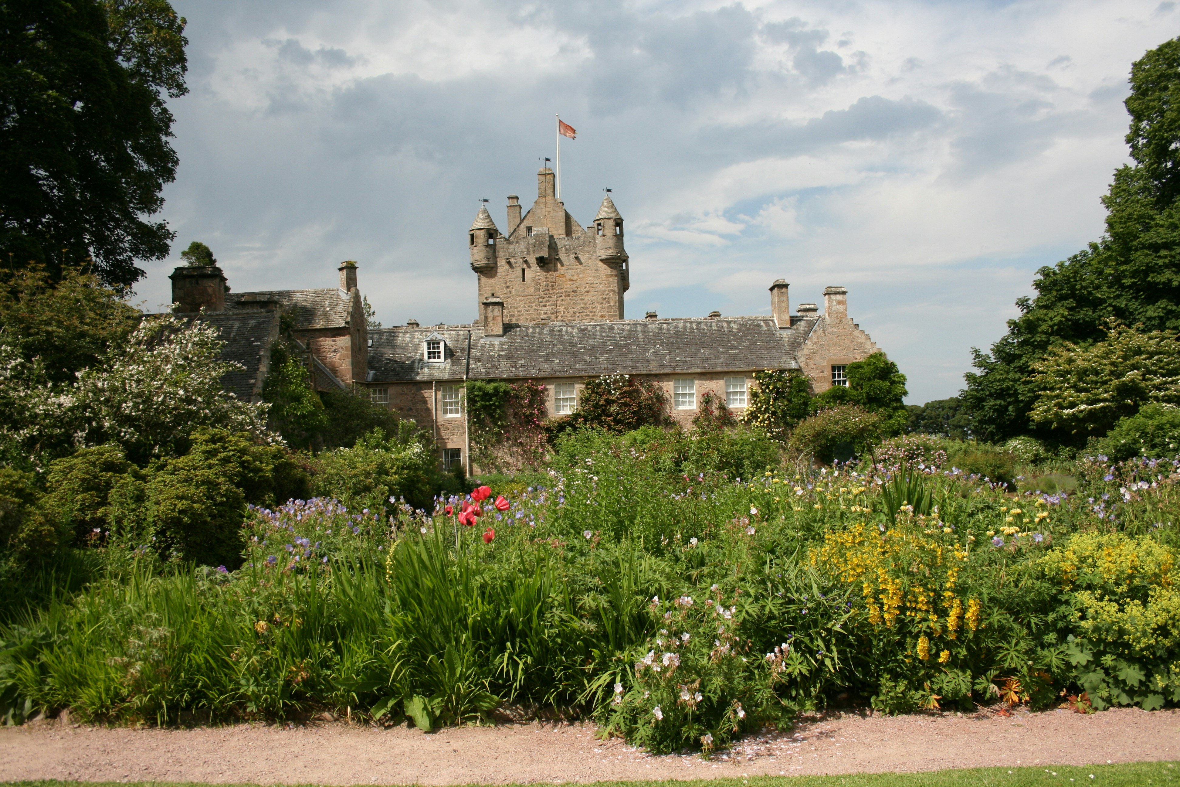 Victorian-era castle surrounded by a vibrant garden filled with colorful flowers and lush greenery. A flag flutters atop the castle tower.