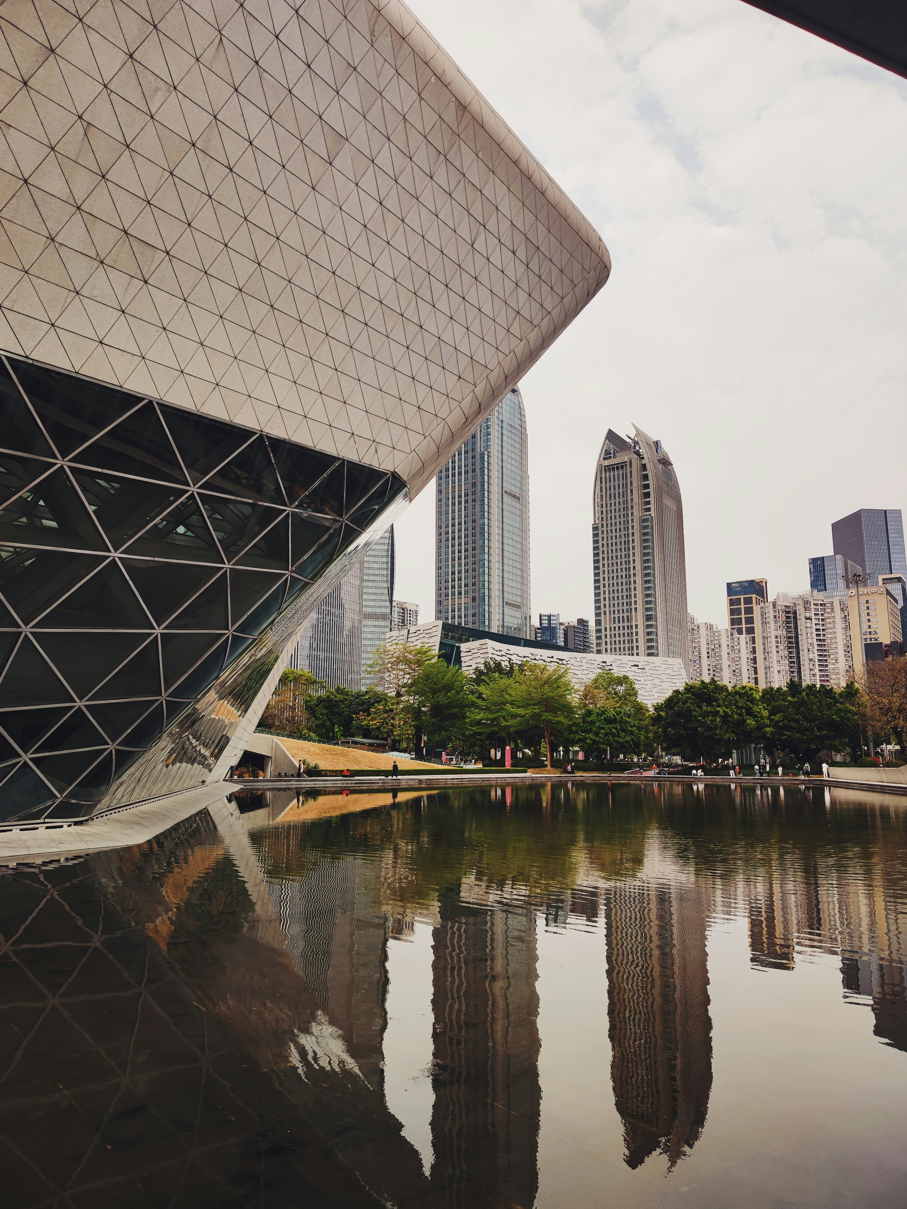 Contemporary building with a geometric facade reflecting in a tranquil pond, surrounded by urban greenery and skyscrapers.