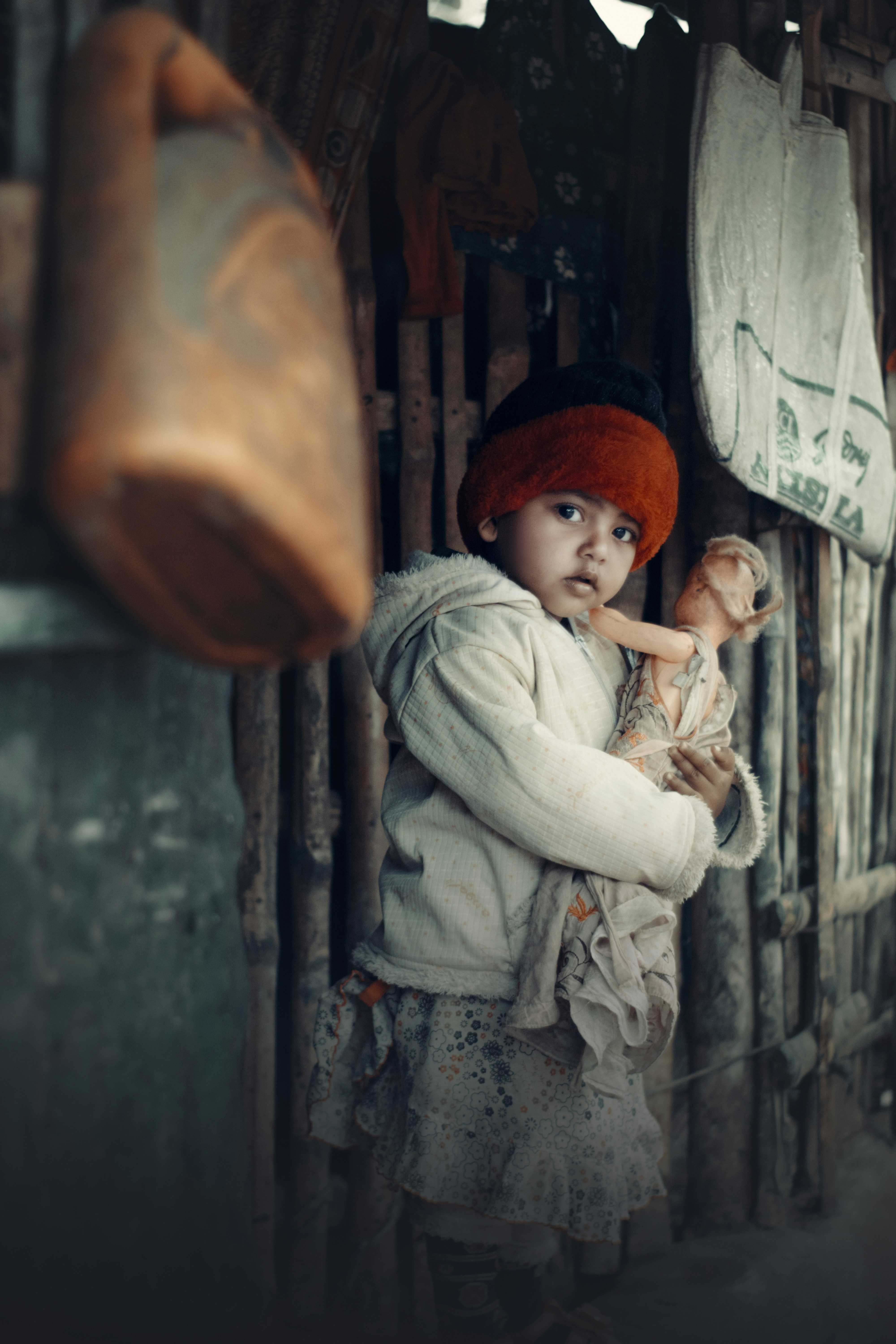 Cornered Warmth in the Alley: A Child in an Orange Hat Clutches a Doll
