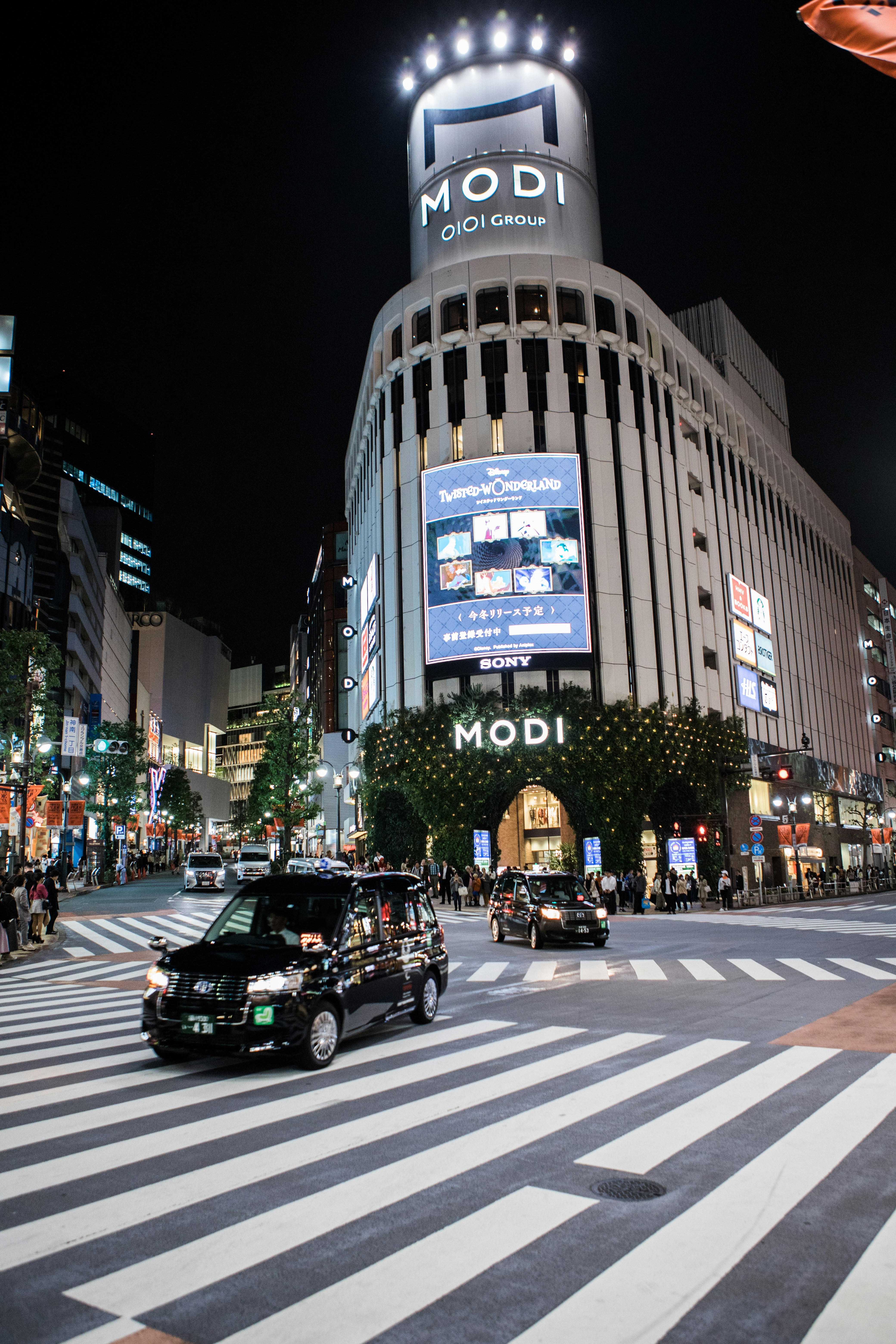 Nighttime photograph of Ginza's MODI building with illuminated signage towering over a busy zebra-striped crosswalk.