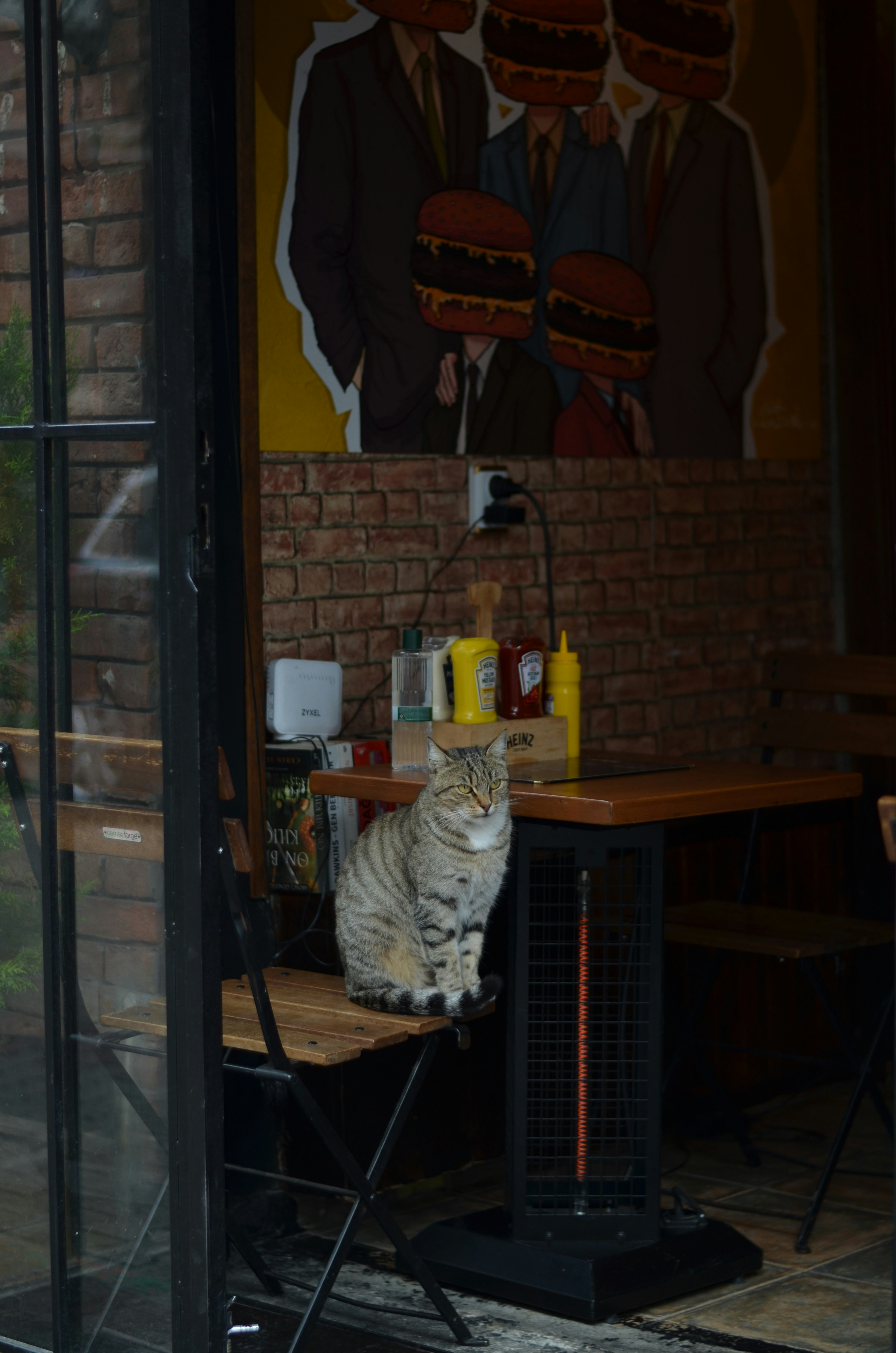 A tabby cat perched on a chair in a cozy café, surrounded by condiment bottles and a quirky hamburger-themed mural. The warm ambiance invites relaxation.