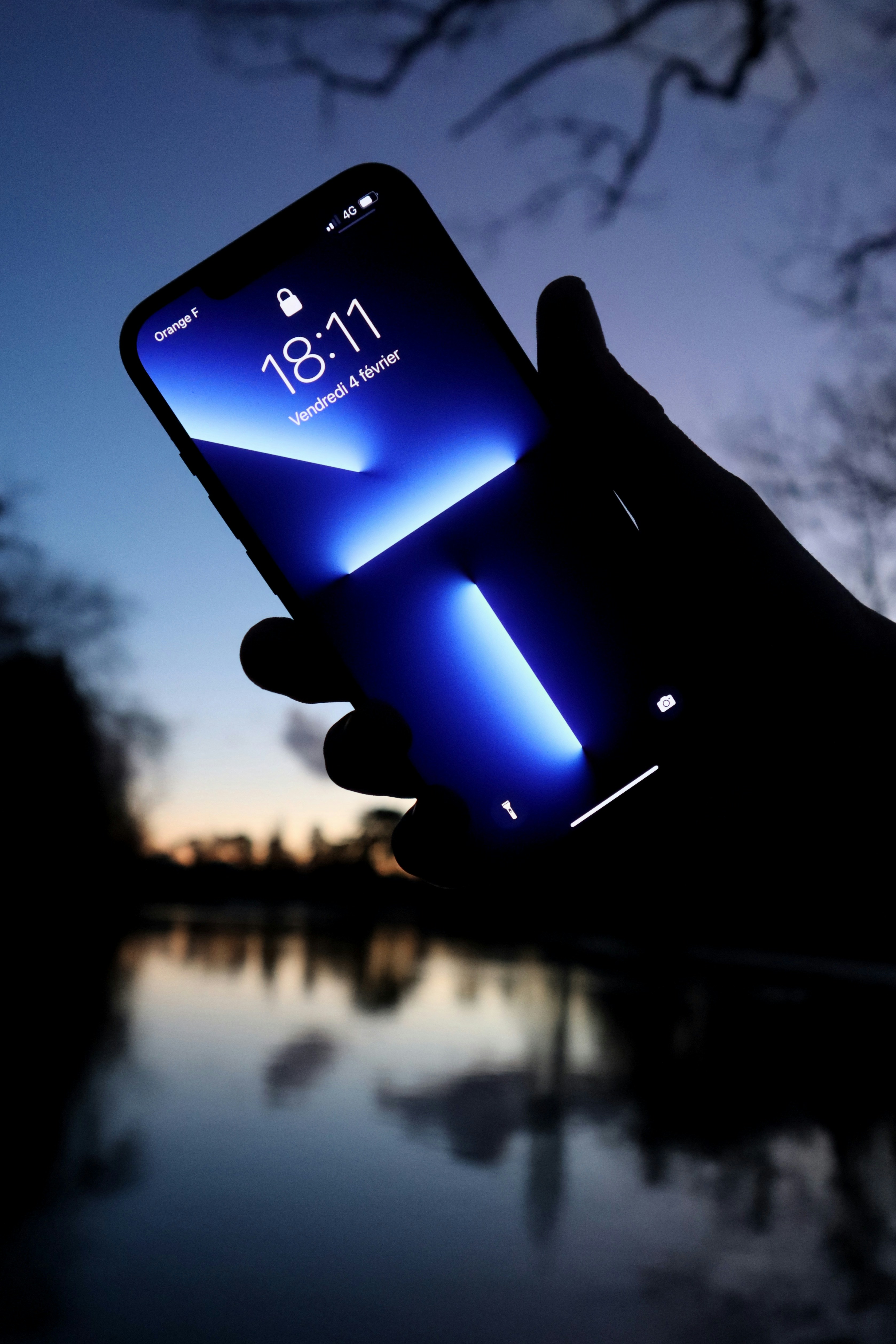 A smartphone displaying the time at dusk, held against a tranquil water backdrop reflecting the fading light. 