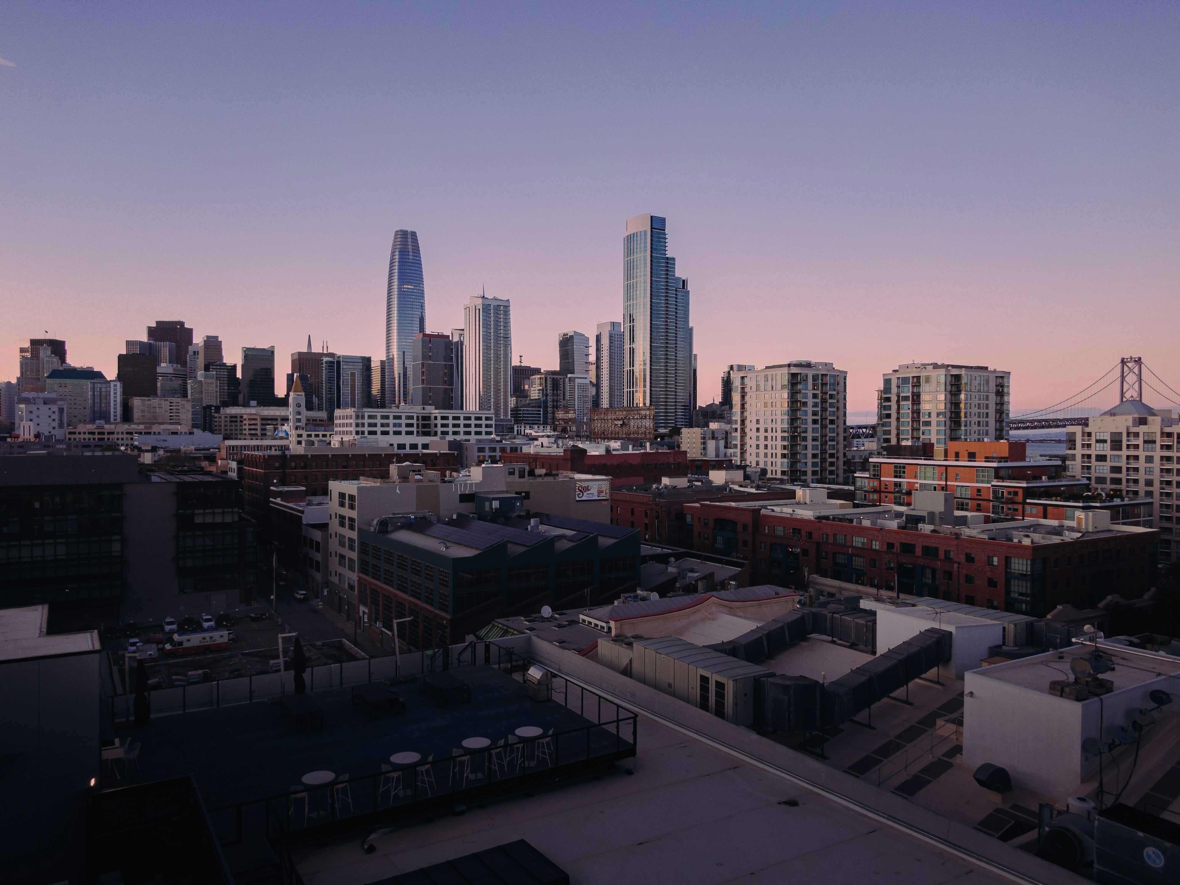 City skyline featuring modern skyscrapers illuminated by twilight, with the Bay Bridge visible in the background.