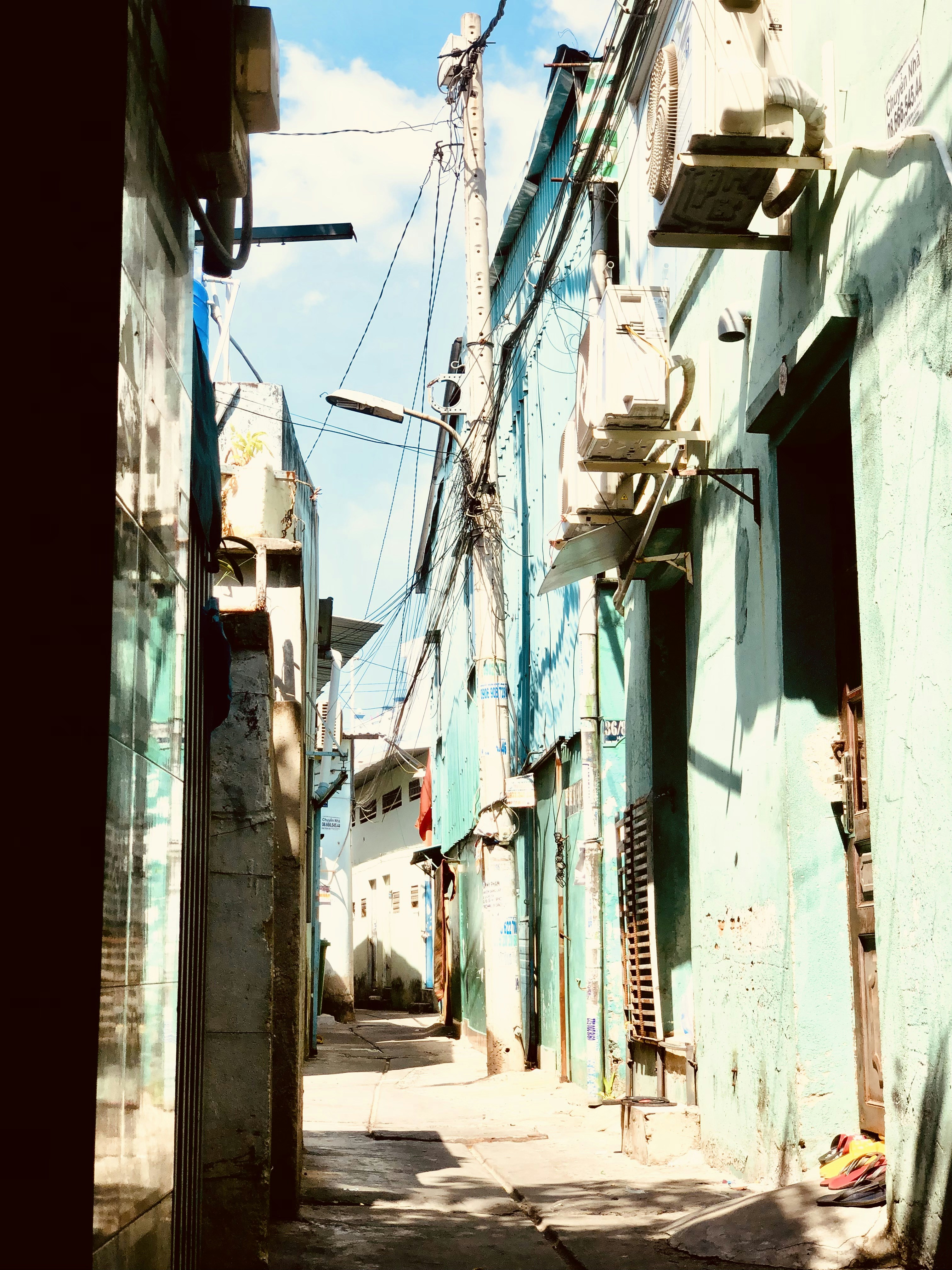Narrow alleyway lined with vibrant teal buildings and air conditioning units, illuminated by soft sunlight. The scene captures a tranquil moment in an urban setting.