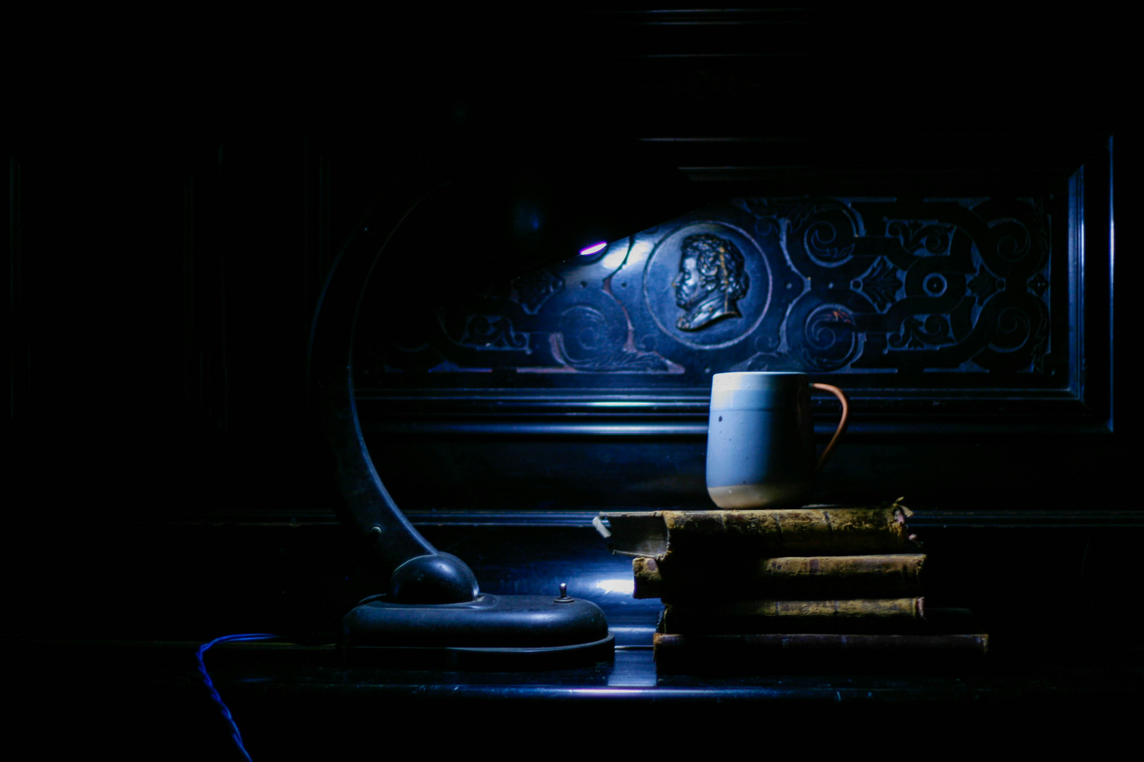 A desk lamp casts a soft glow on a stack of vintage books beside a ceramic mug, creating a tranquil atmosphere in a dimly lit setting.
