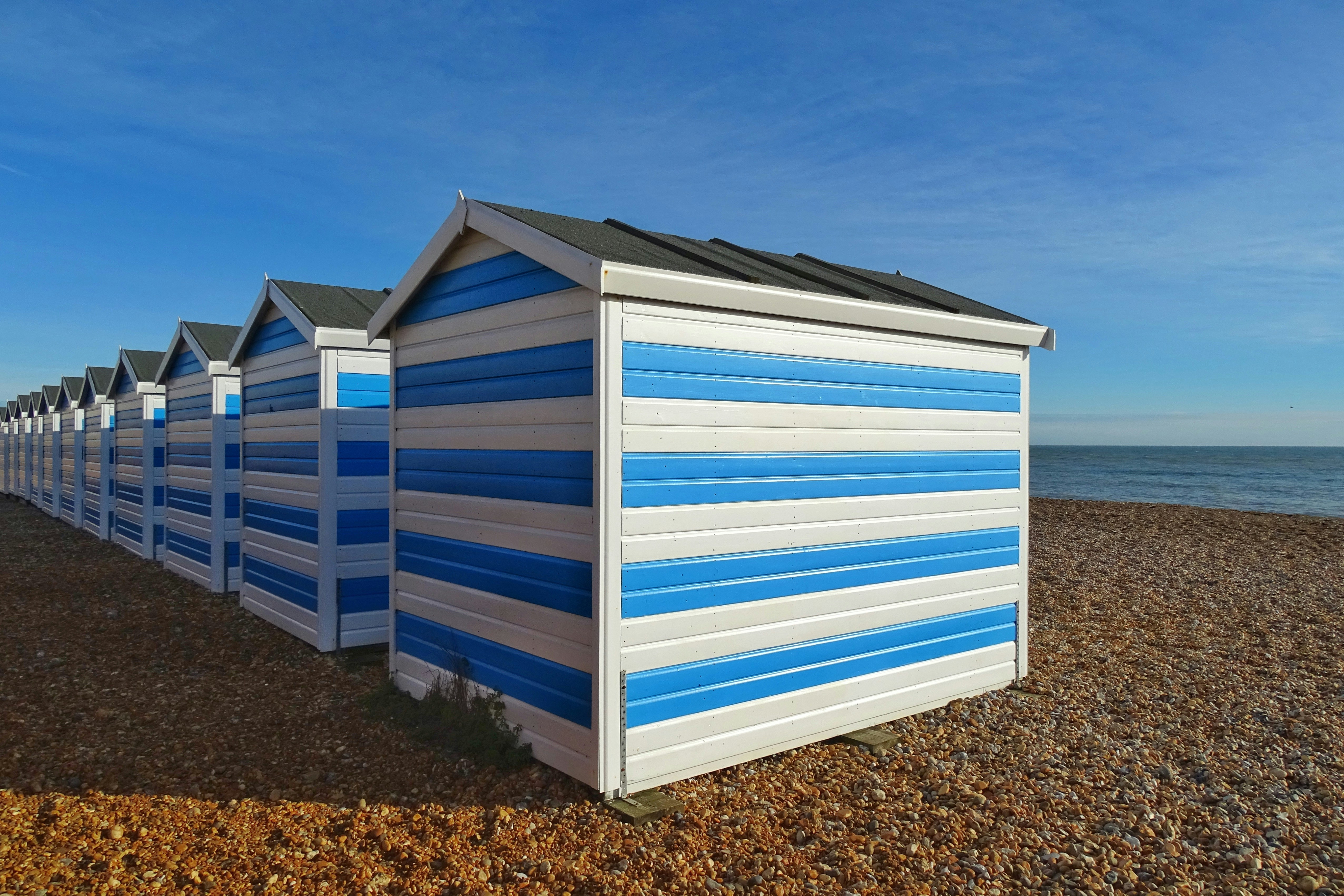 Stripe-by-stripe procession: blue beach huts along a pebbled shoreline
