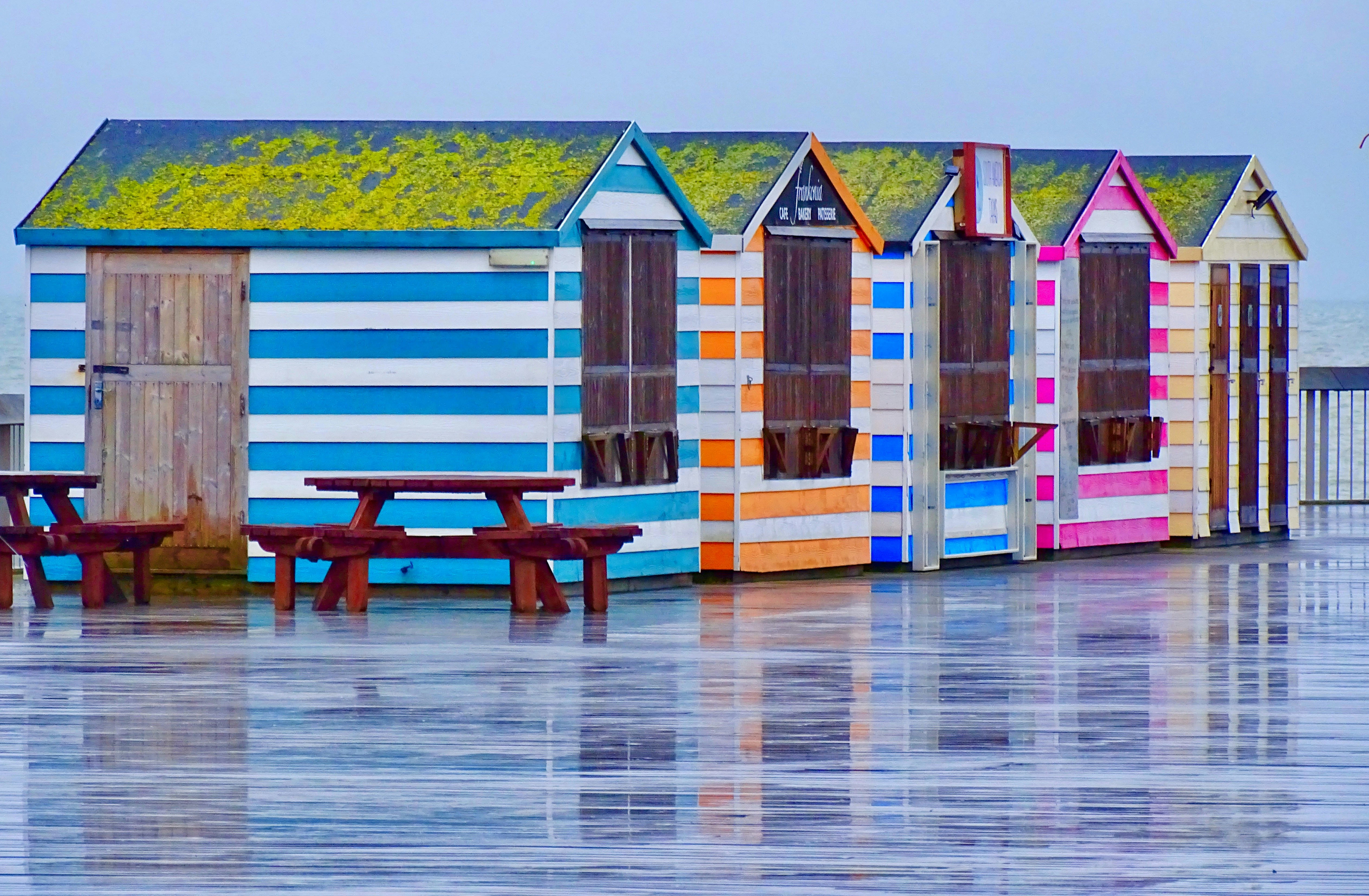 Colorful beach huts line Hastings Pier, their vivid stripes mirrored on the wet surface.