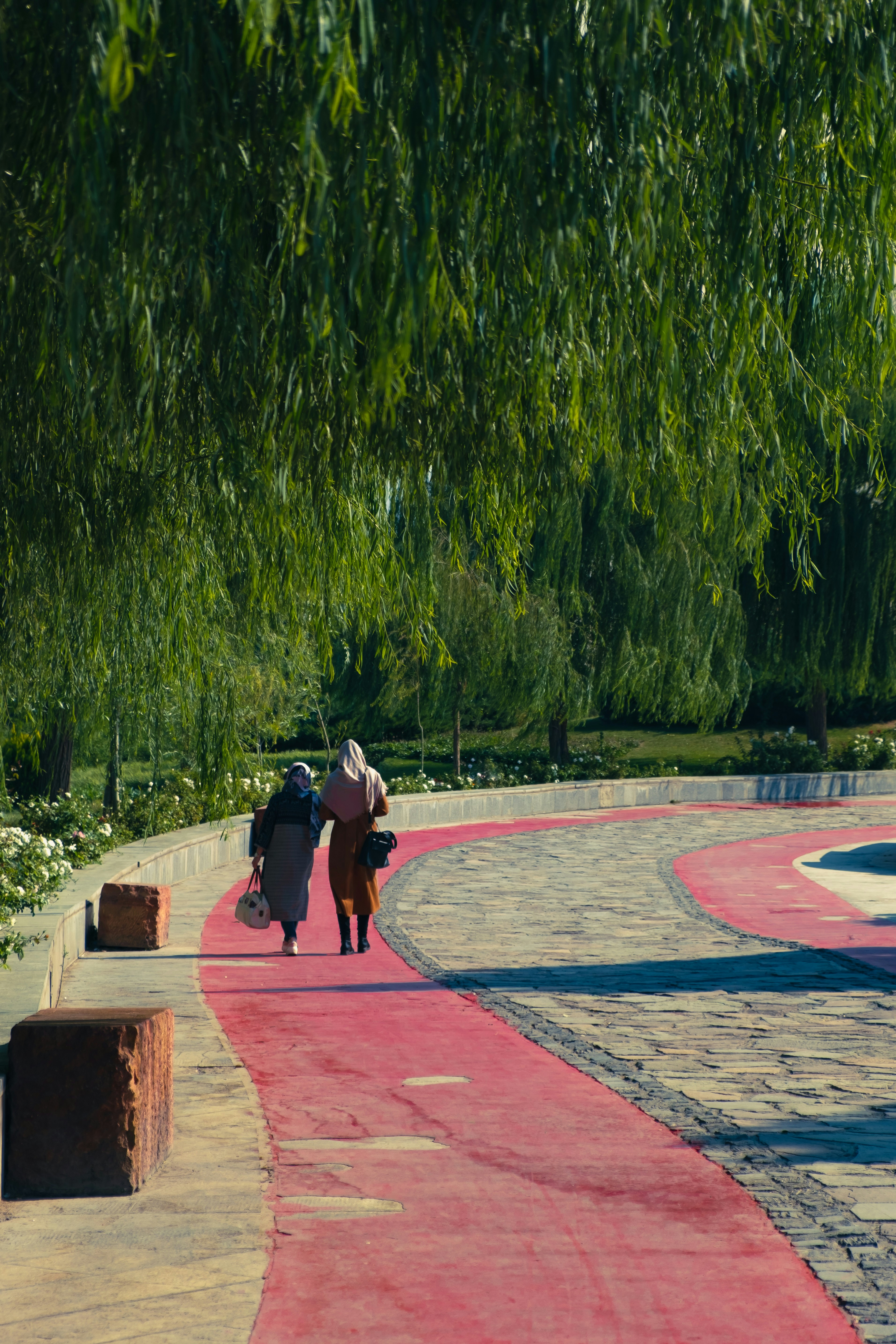 Two women stroll along a curving pathway adorned with vibrant colors, flanked by lush greenery and willows. The scene conveys a sense of tranquility and companionship.