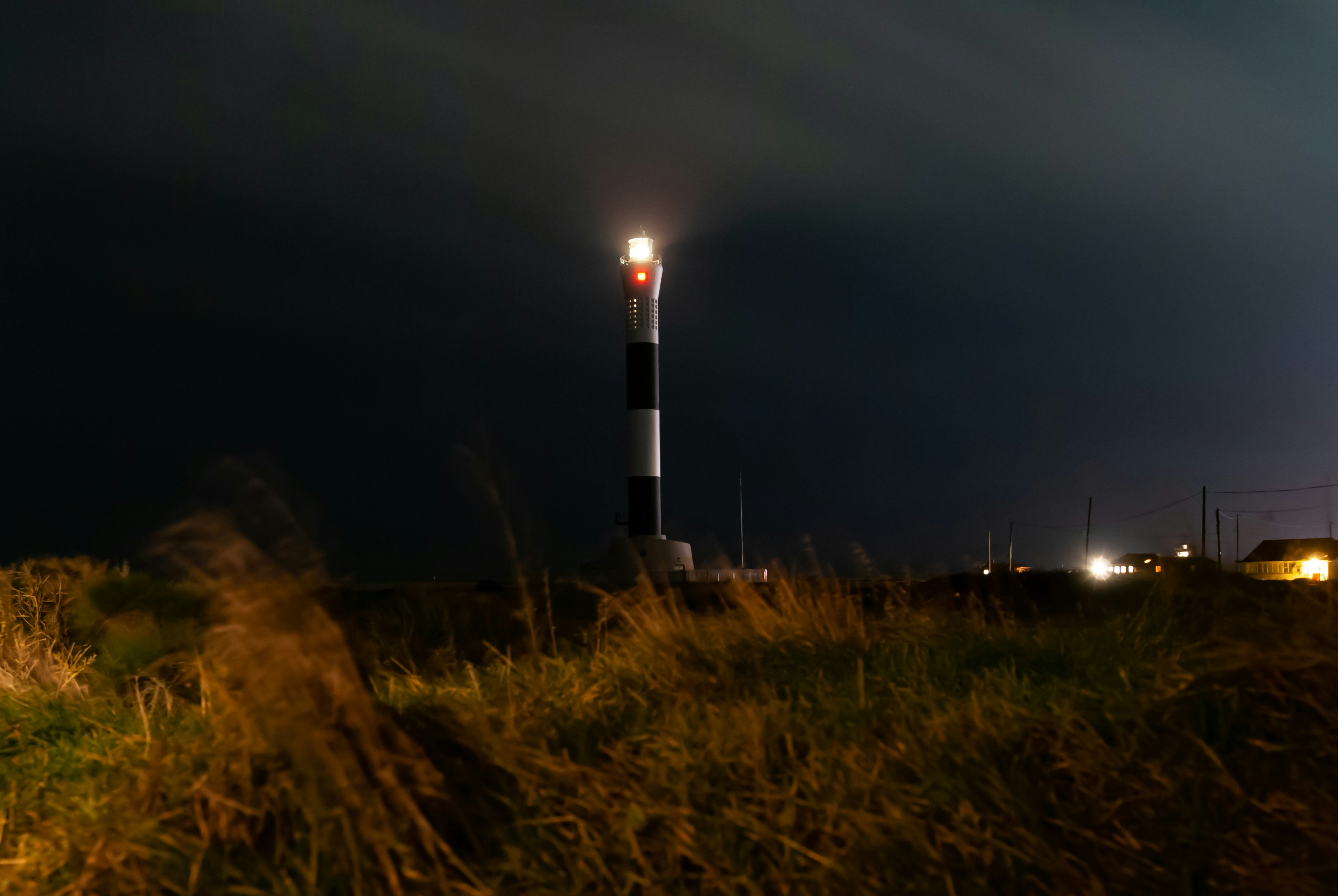 A lighthouse stands tall against the night sky, its light piercing through the darkness while surrounding grass sways in the breeze.