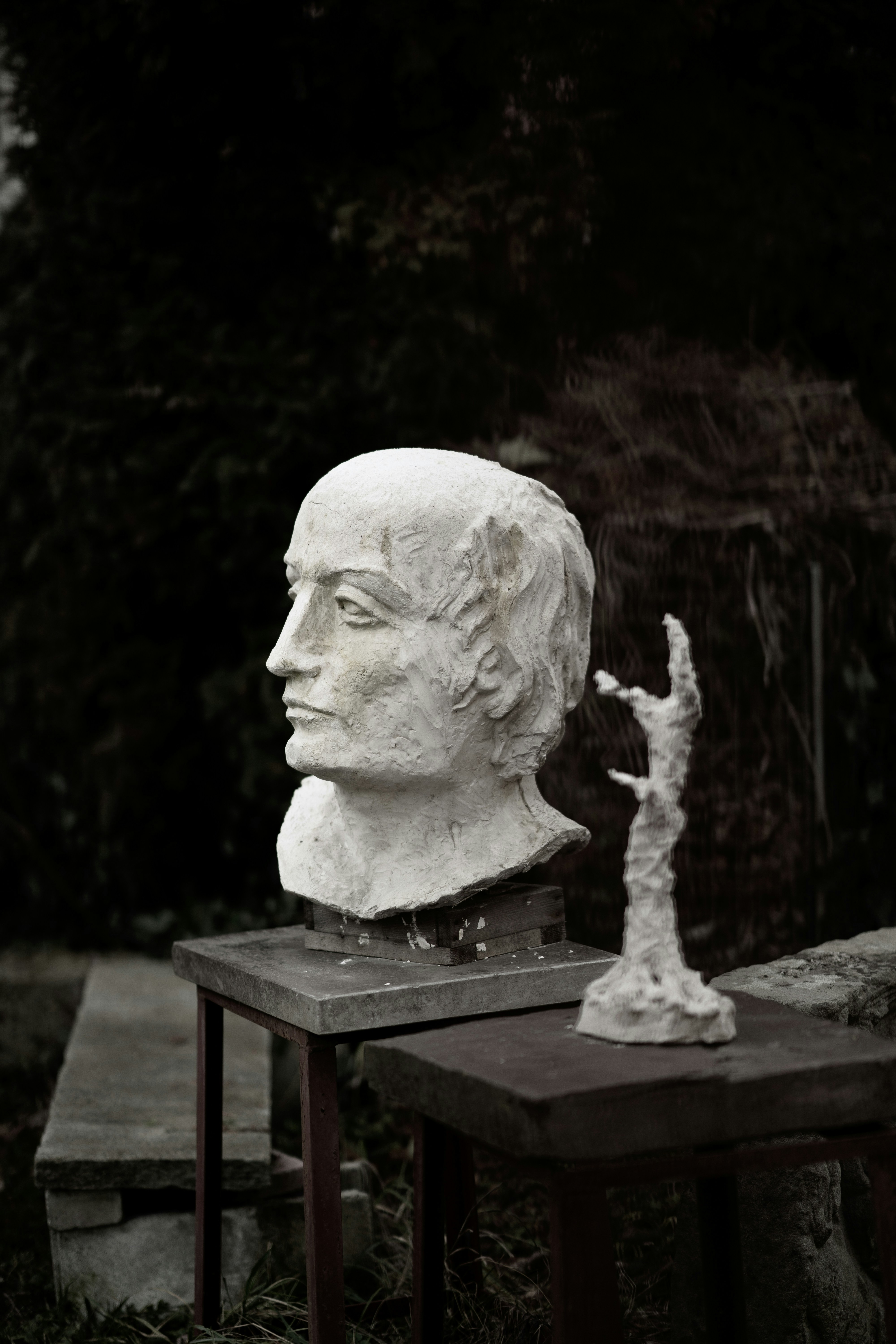 A white sculpted bust and a smaller figure on wooden tables, set against a muted background, showcasing the intricacies of artistry in a serene outdoor setting.