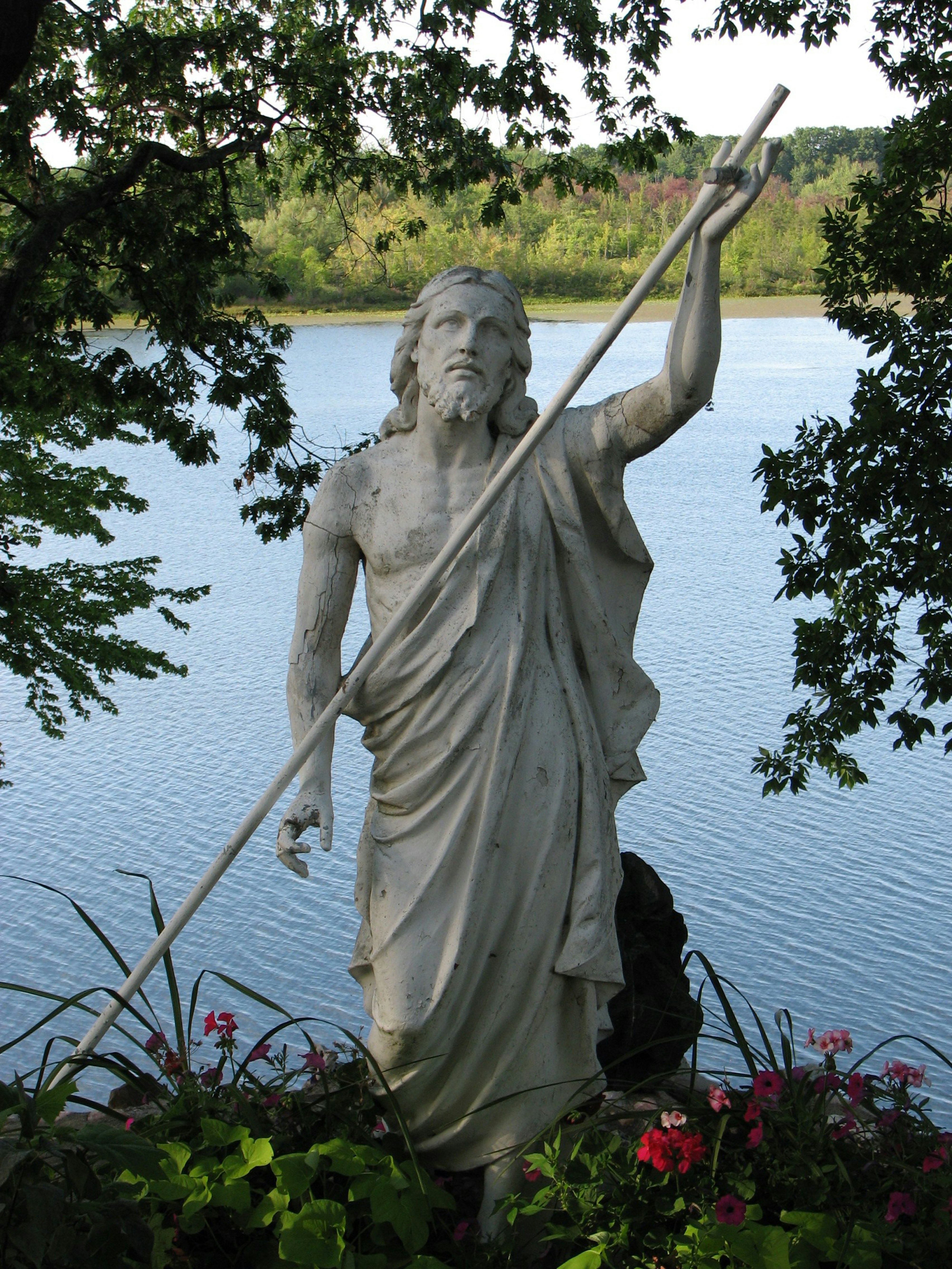Marble statue of a draped figure holding a spear stands beside a calm lake, framed by trees and blooming flowers.