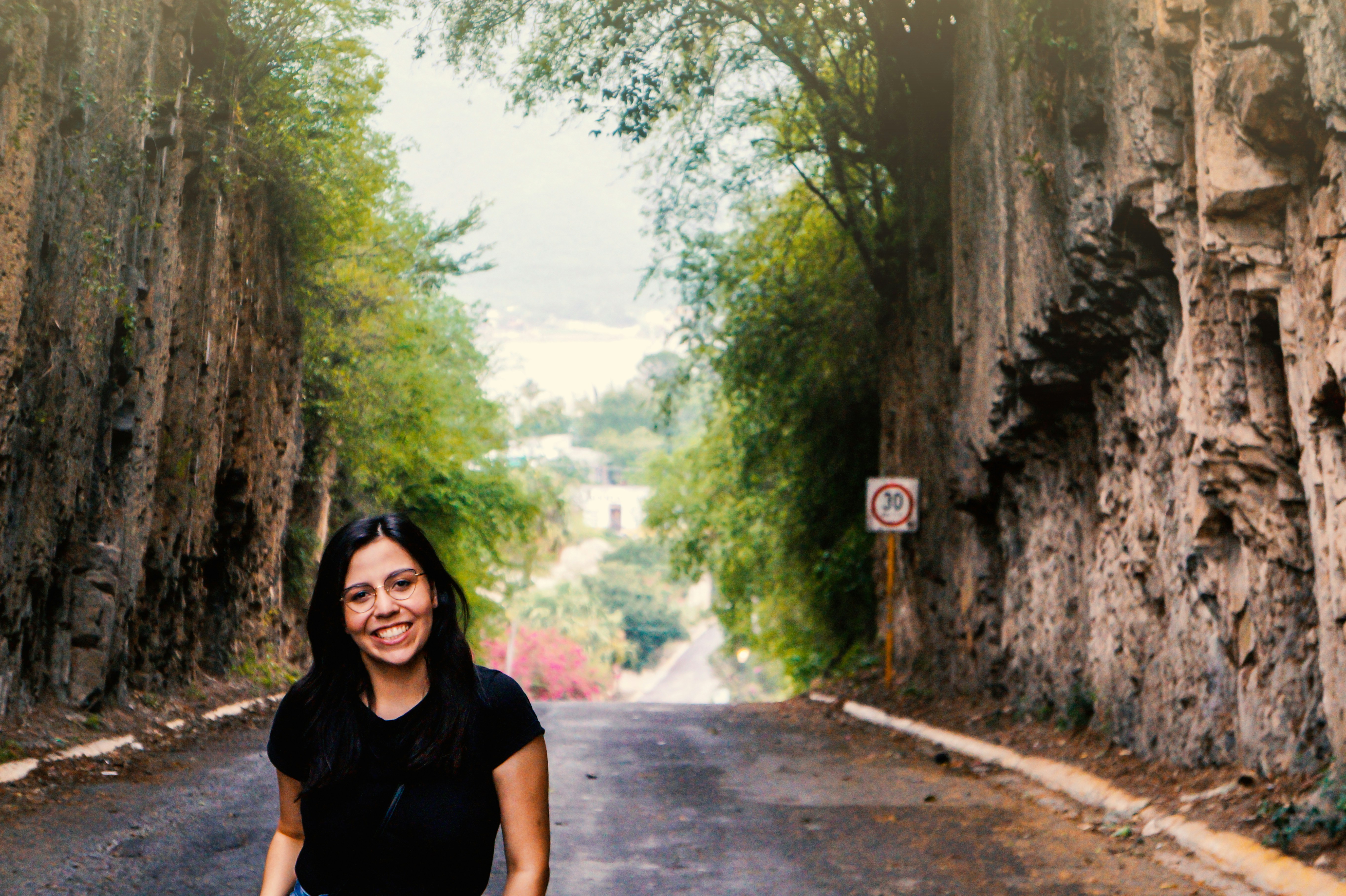 Woman smiling on a road flanked by steep, rocky cliffs and lush greenery.