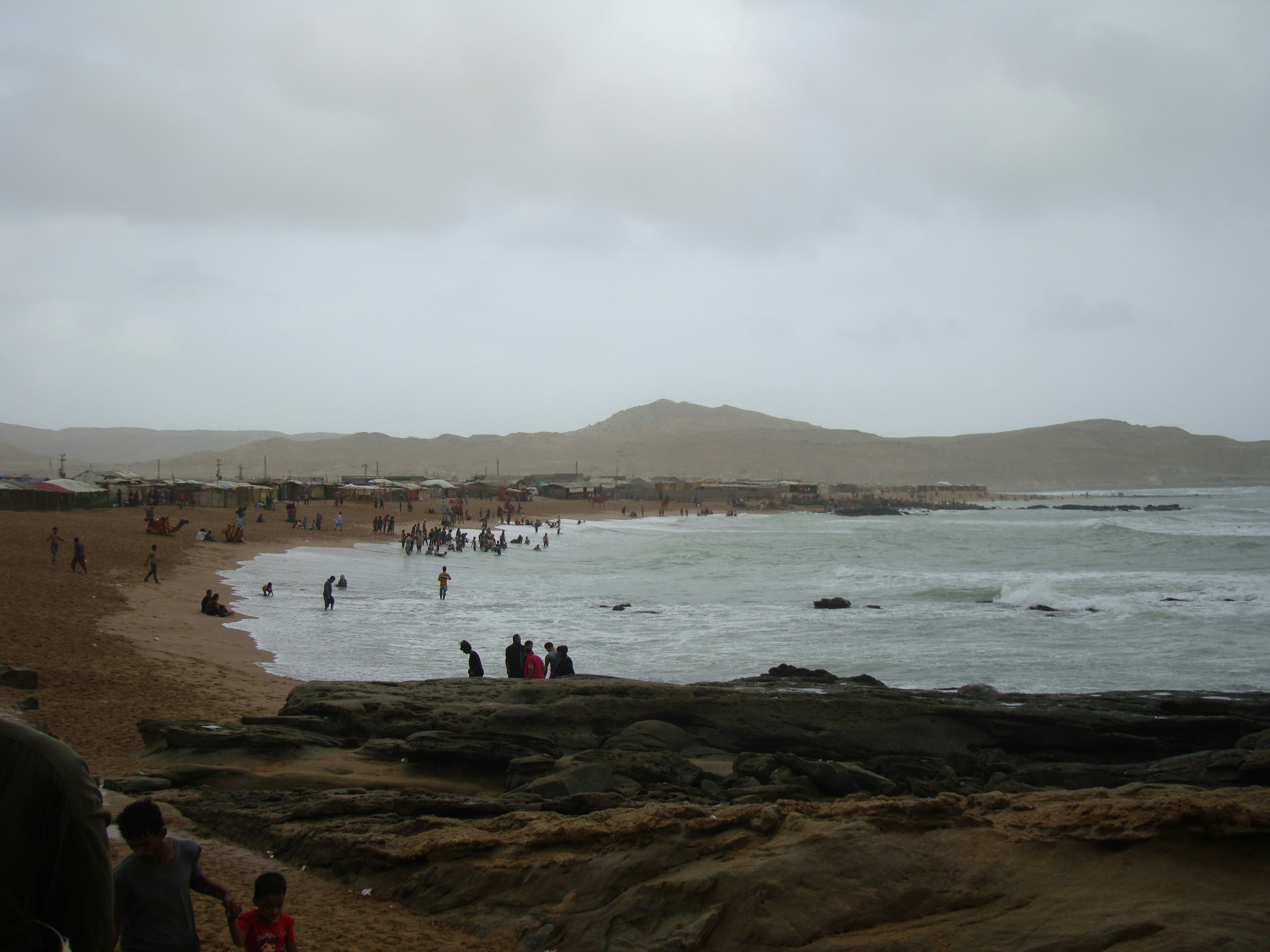 Crowds of beachgoers enjoy a cloudy day at the shoreline, with rocky formations contrasting the sandy beach and gentle waves. 