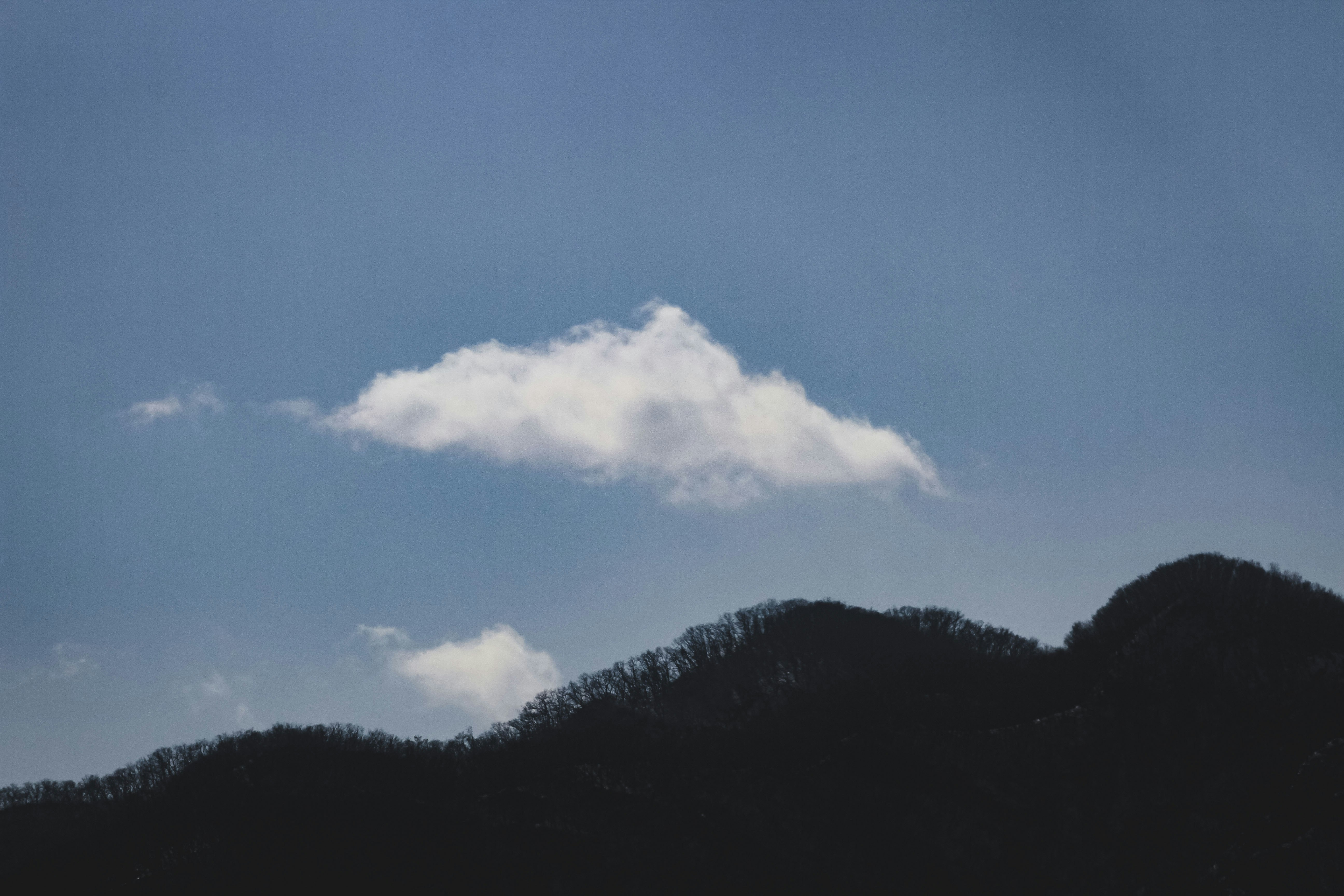 A solitary cloud drifts above a silhouette of rolling hills under a clear blue sky.