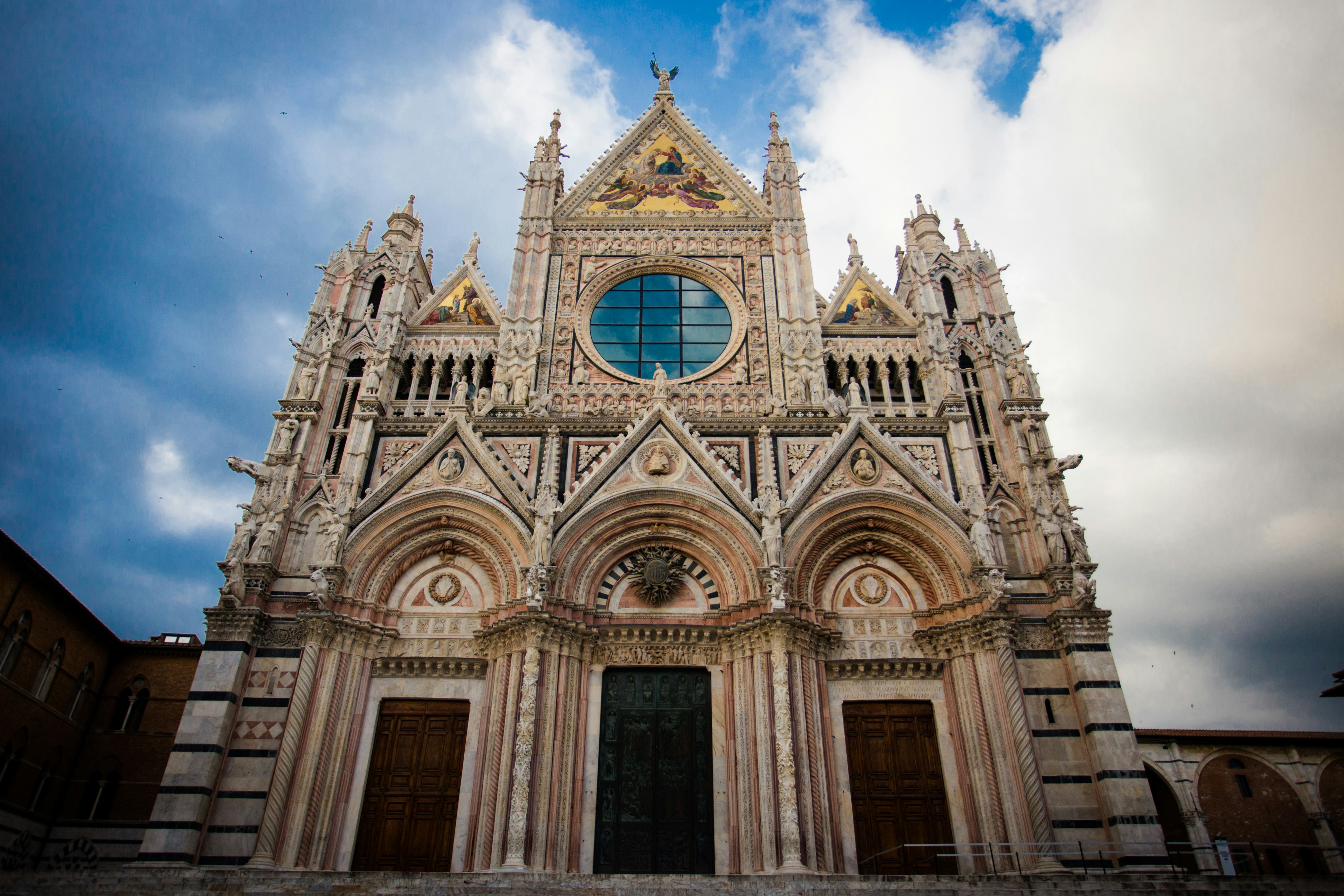 Gothic cathedral facade with intricate stonework and a large circular stained glass window beneath a partly cloudy sky.