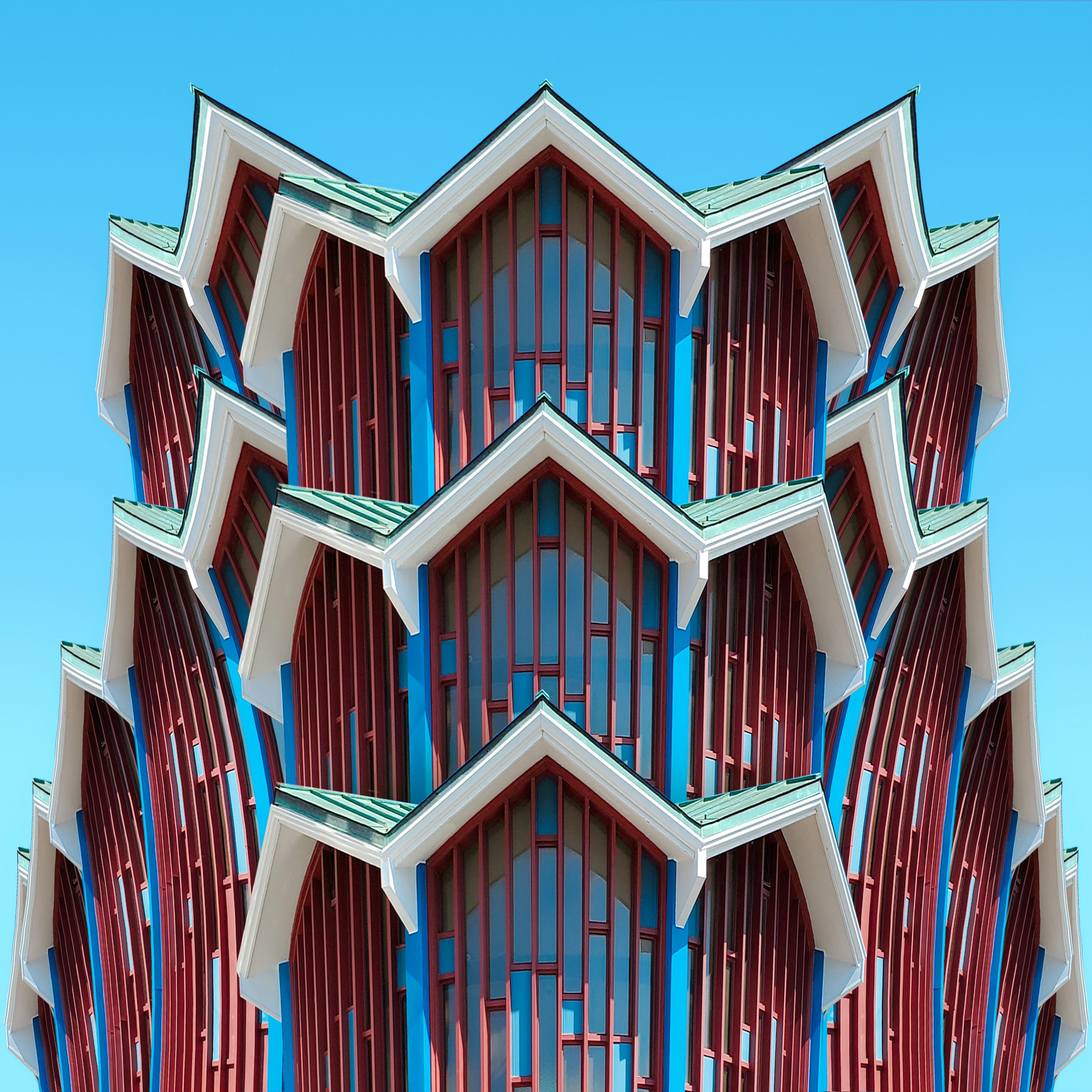 Bold multilevel building facade with repeating red slats and white frames forming chevron-like peaks against a bright blue sky.