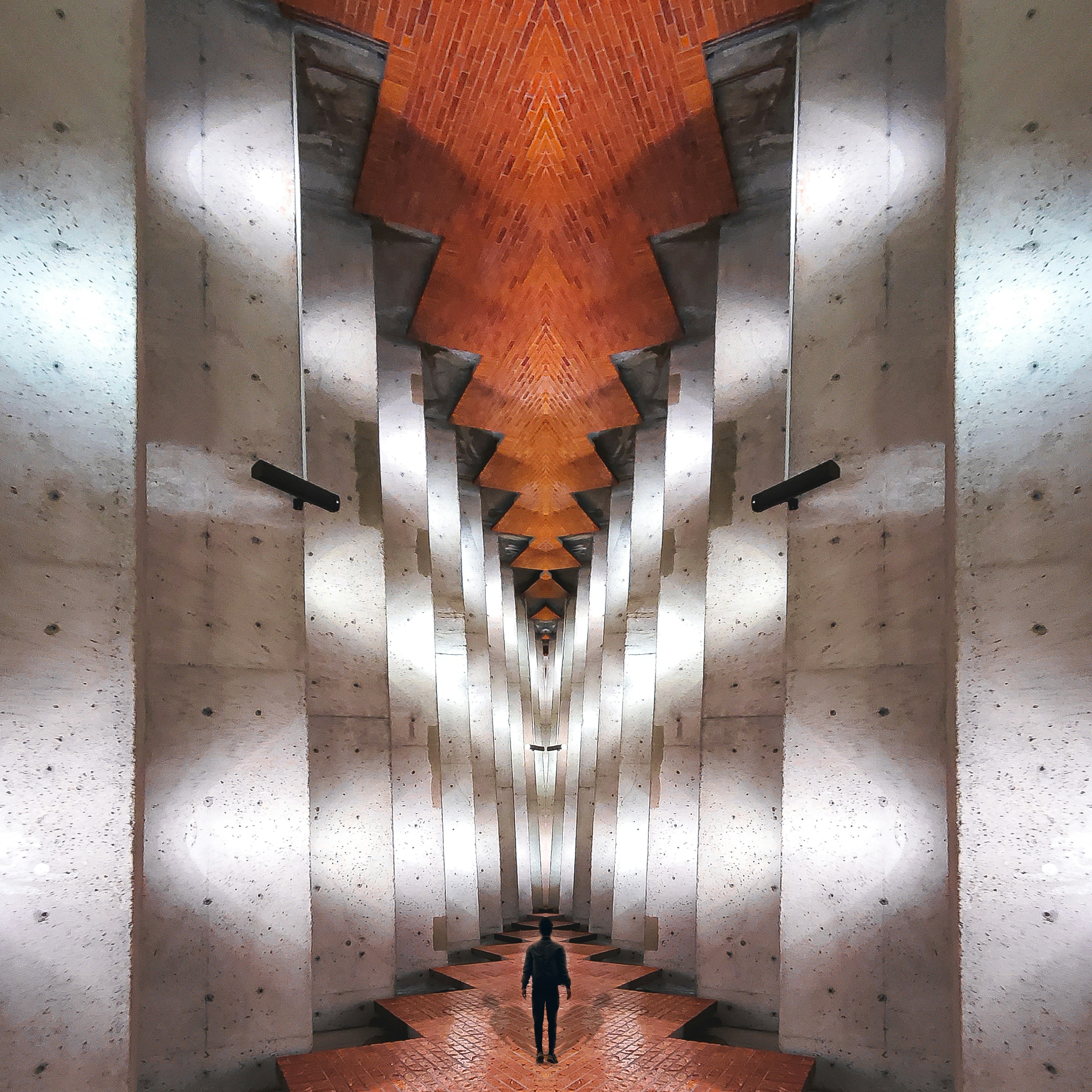 A solitary figure stands at the center of a vast architectural space, surrounded by towering concrete columns and contrasting warm brick ceilings. The interplay of light and shadow creates a mesmerizing depth effect.