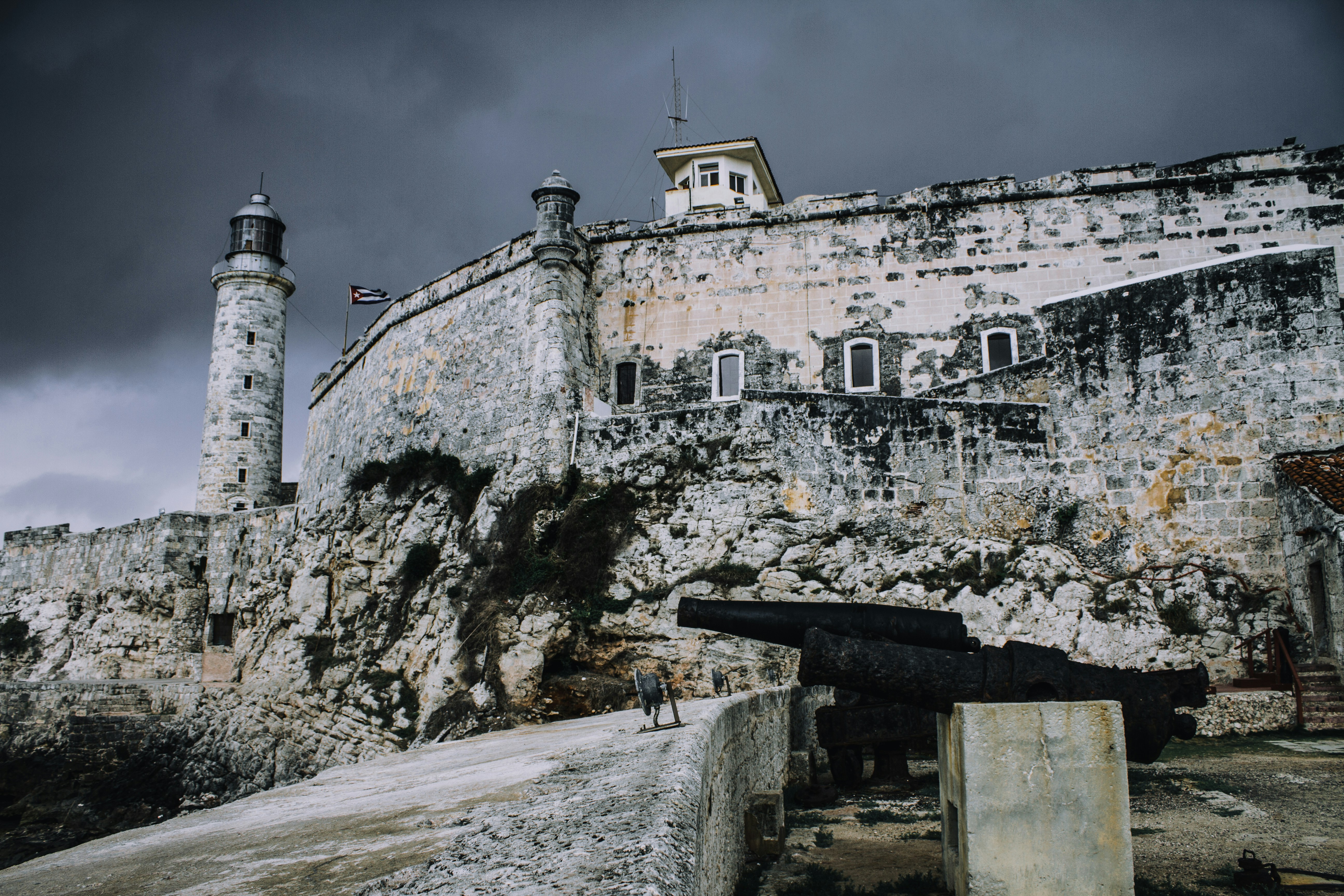 Historic fortress with a lighthouse and cannons overlooking a rocky coastline under a moody sky.