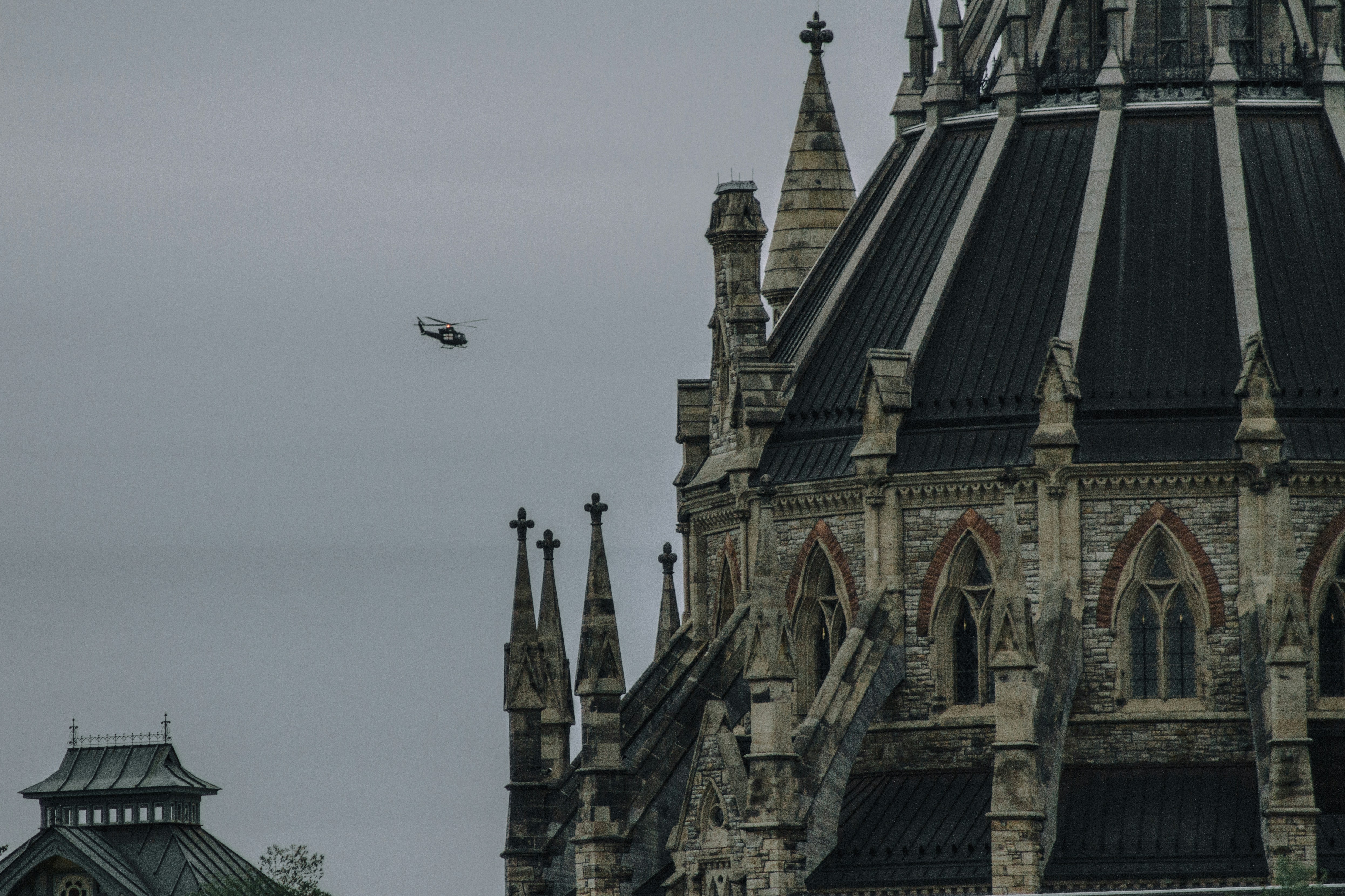 Helicopter hovering near the ornate steeple of a historic cathedral under a cloudy sky.