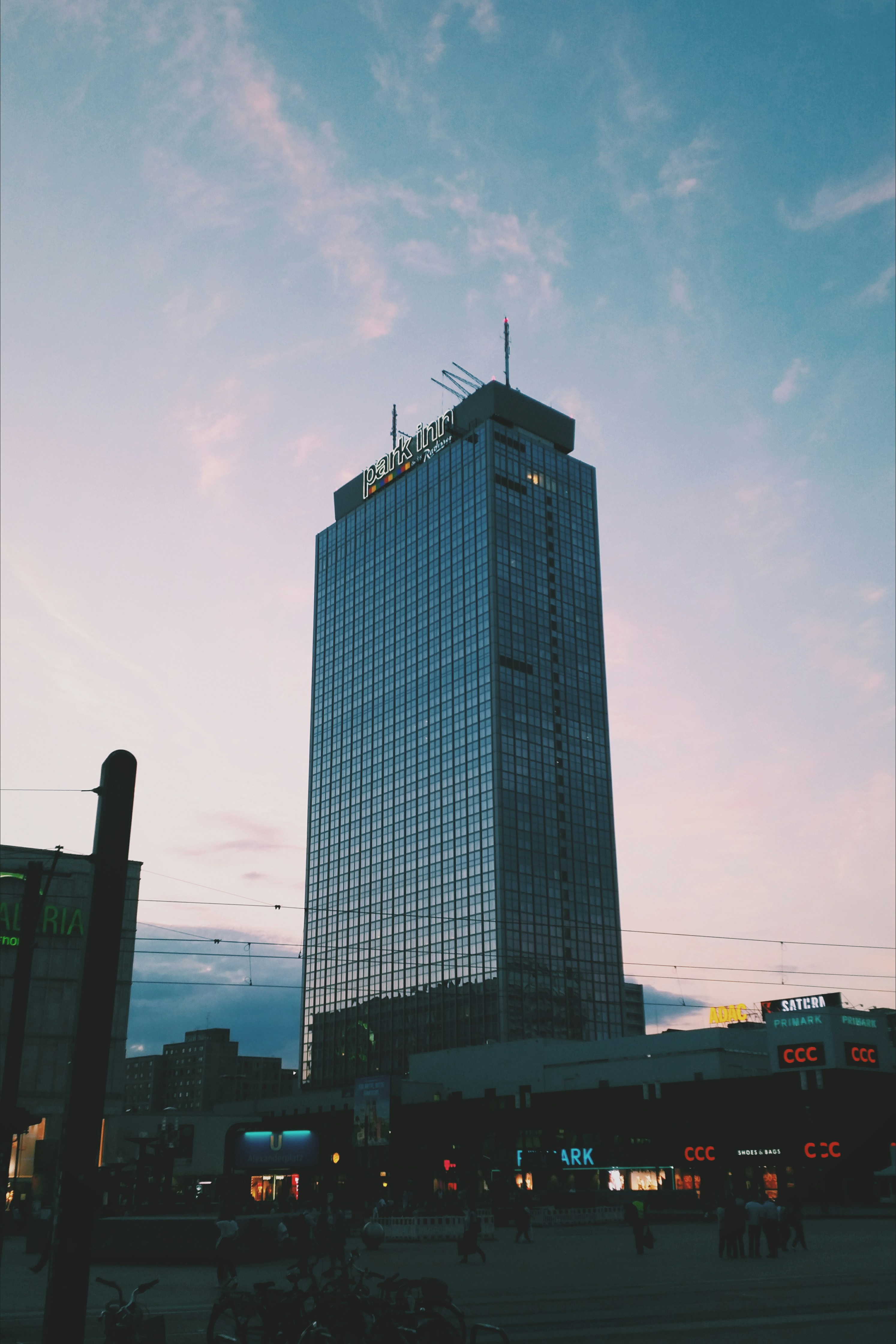 Tall glass skyscraper reflecting the evening sky, surrounded by urban life and vibrant storefronts.