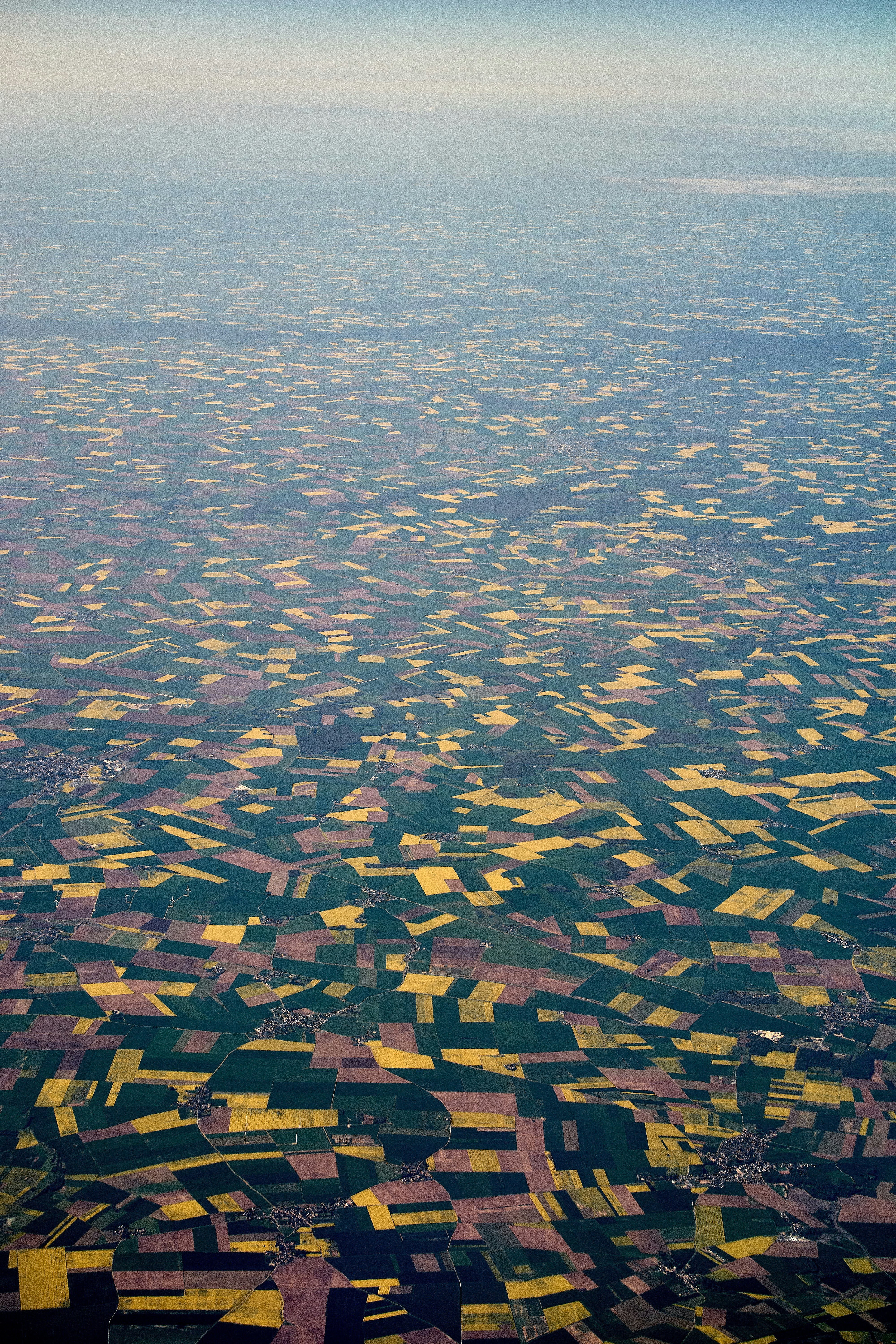 Aerial view of patchwork farmland displaying vibrant green and yellow fields interspersed with brown patches. The landscape reveals the intricate patterns of agricultural land use.