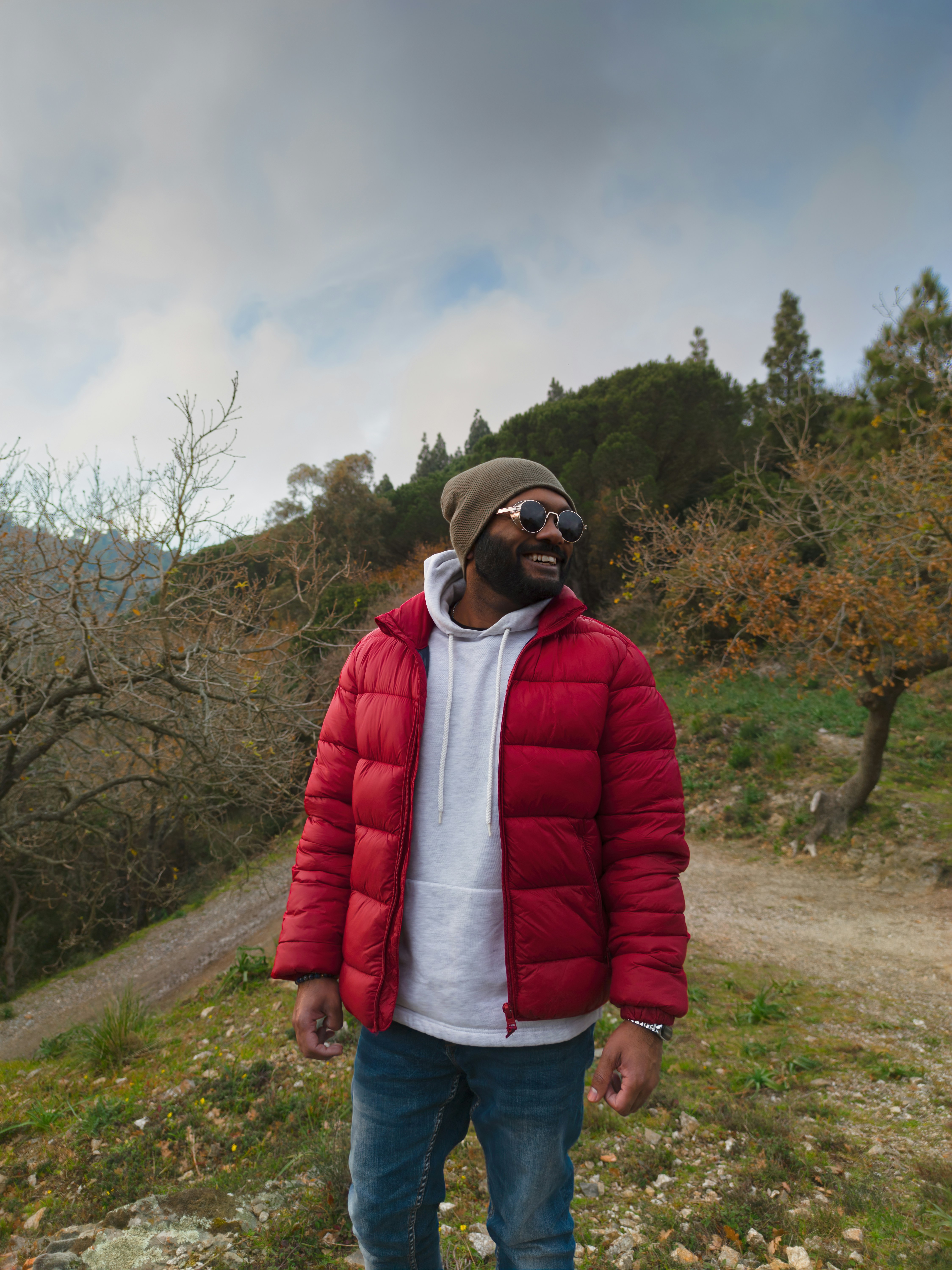 Man in a red puffer jacket and sunglasses stands on a winding path, surrounded by lush greenery and trees. The scene captures a moment of joy amidst nature.