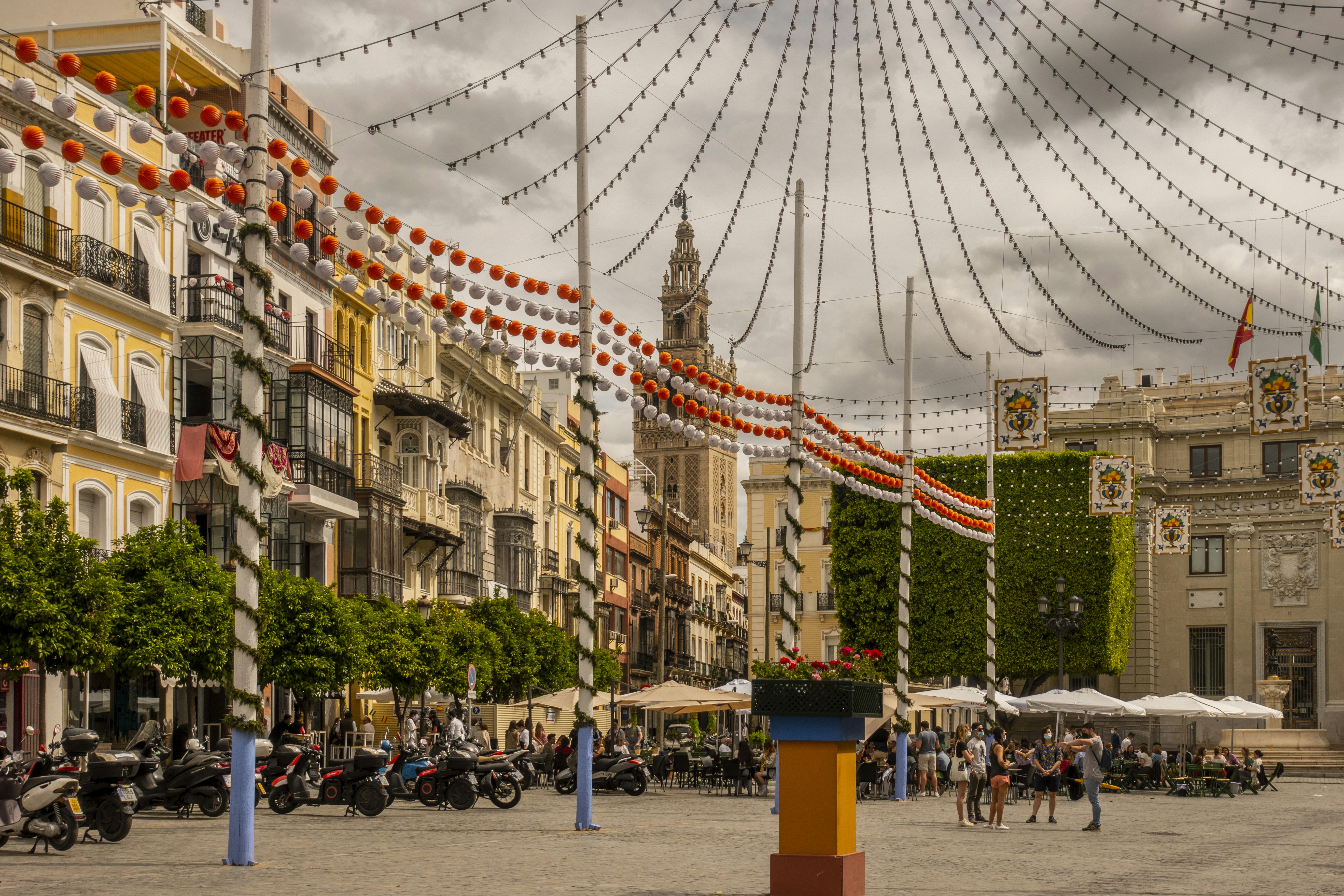 Colorful flags draped across a lively plaza with historic architecture under a cloudy sky.