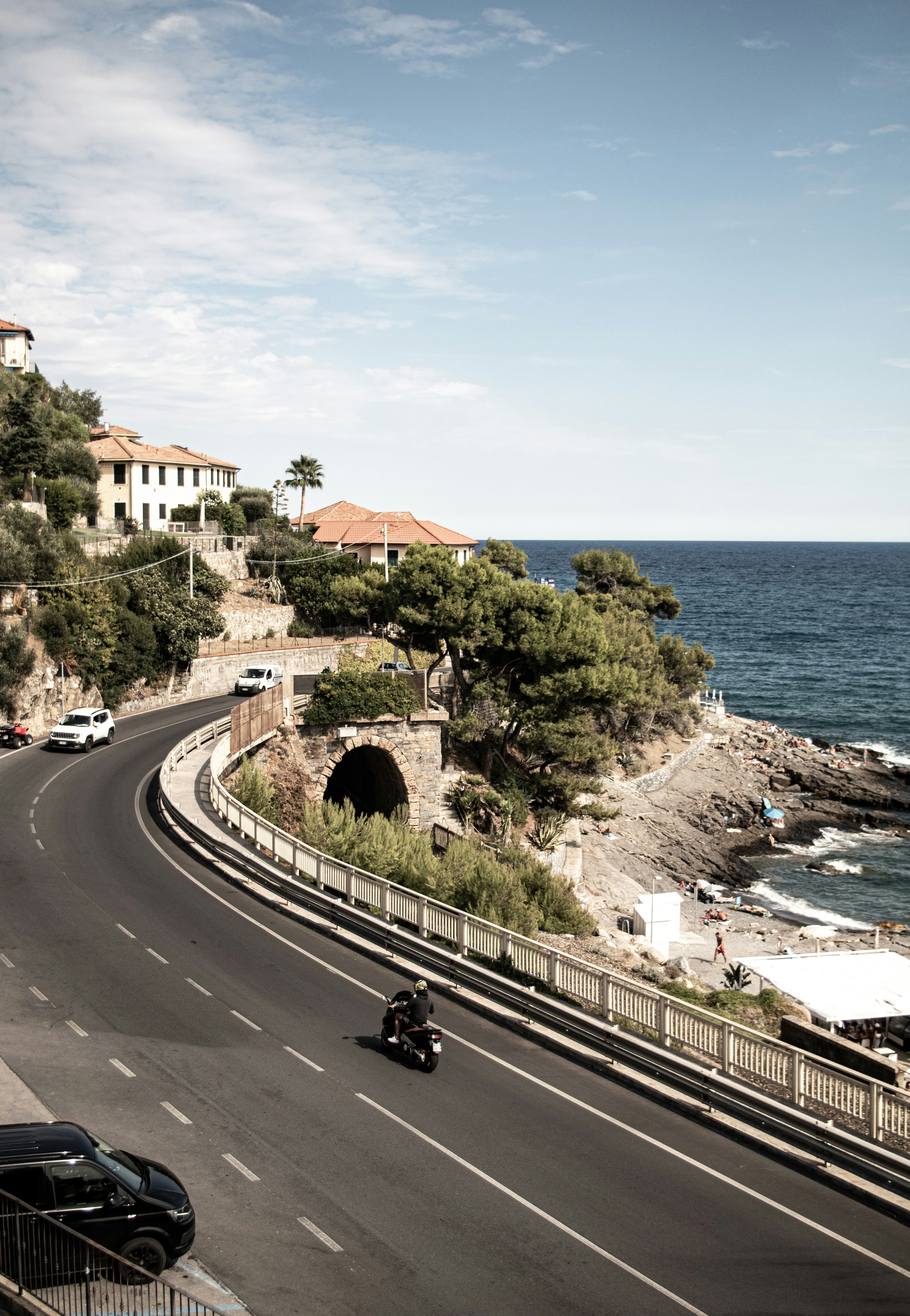 Winding coastal road alongside the sea, framed by lush greenery and charming homes. A motorcycle navigates the curve, evoking a sense of adventure.