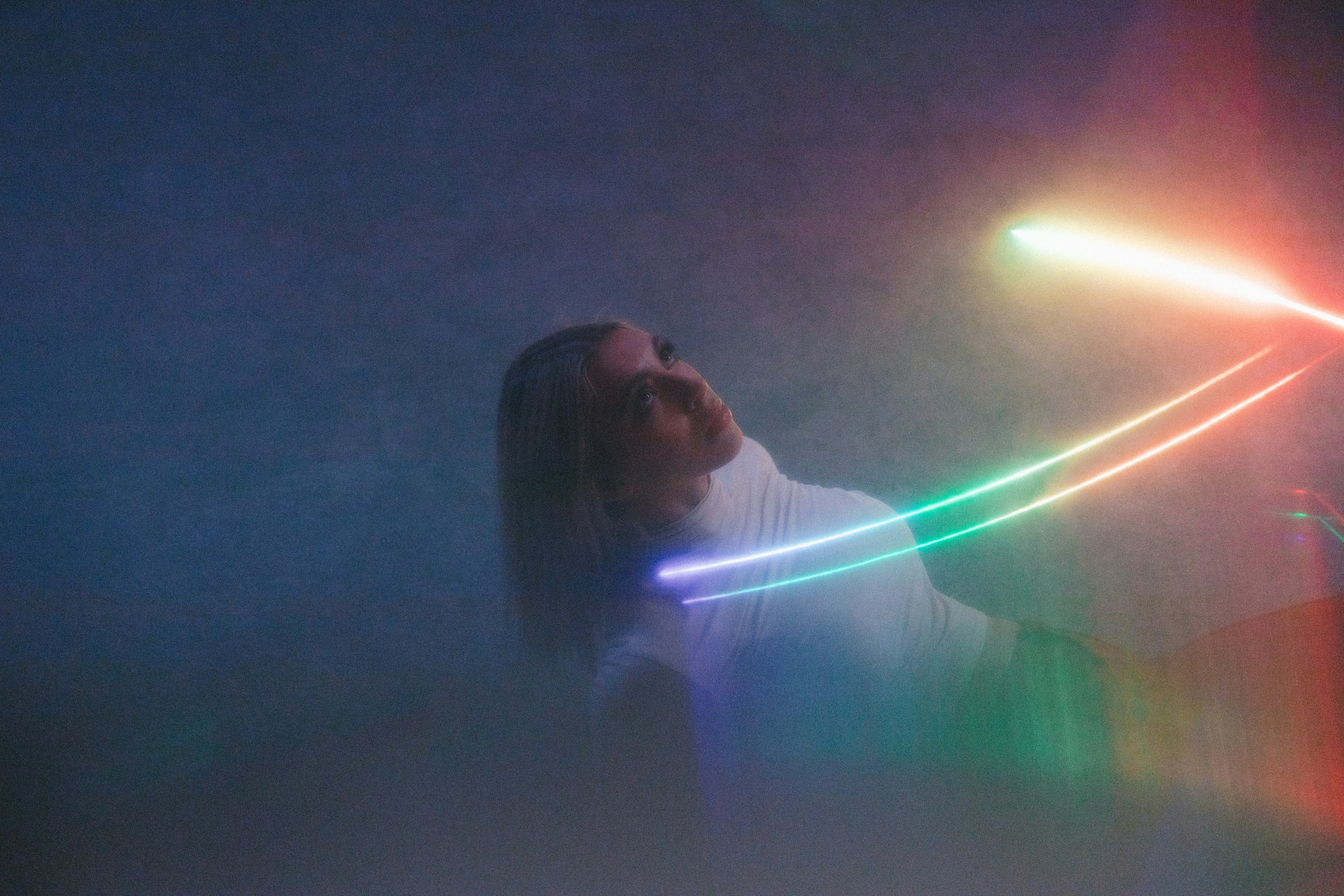 This captivating image features a person gazing upwards, enveloped in an ethereal glow of neon light trails. The composition artfully combines soft, hazy textures with vibrant streaks of color, including blues, greens, and reds, creating a surreal atmosphere. The play of light against the subject's white attire enhances the dreamlike quality, making the image visually striking and immersive.