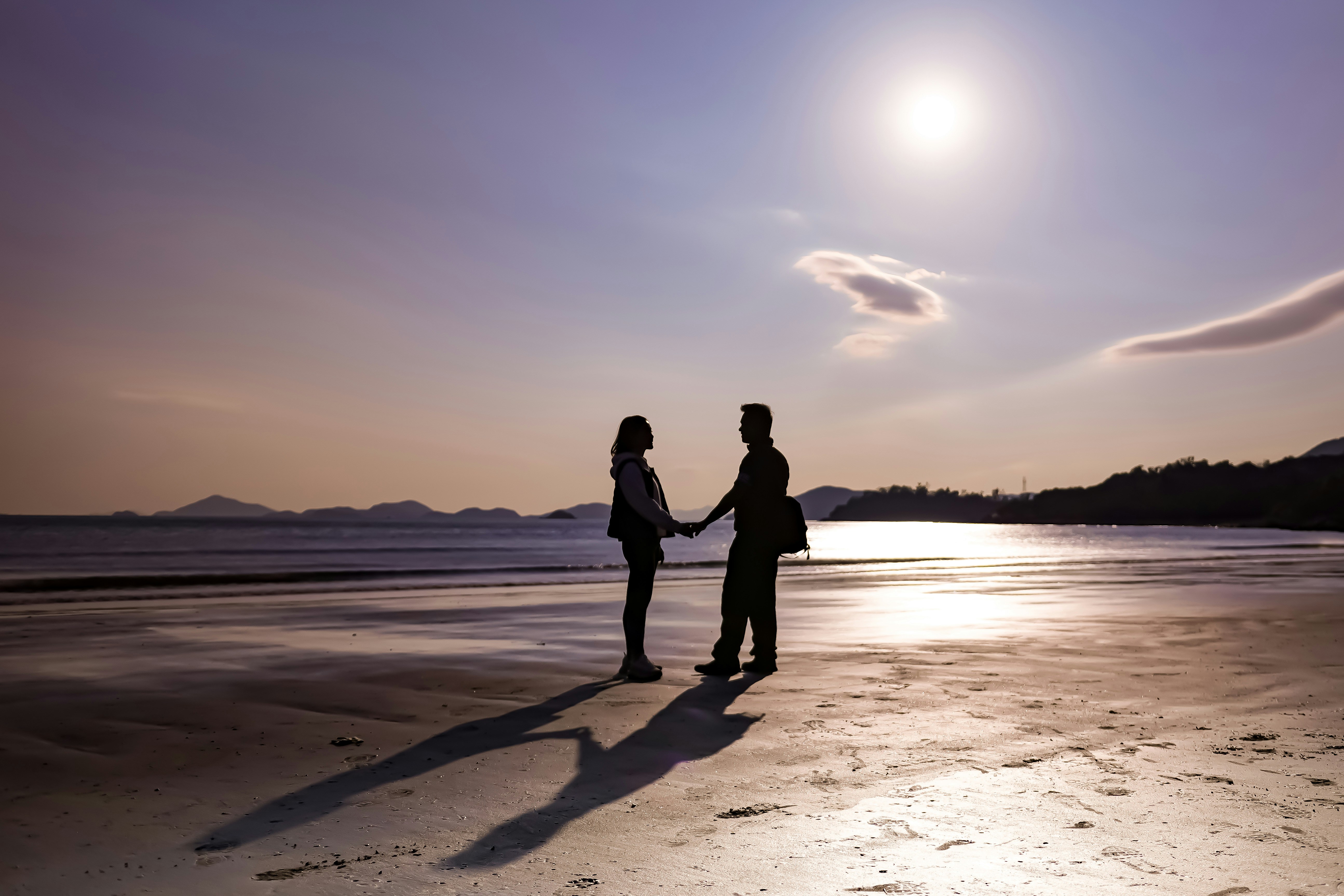 Silhouetted couple holding hands against a serene beach backdrop during twilight, with soft hues reflecting on the water's surface.