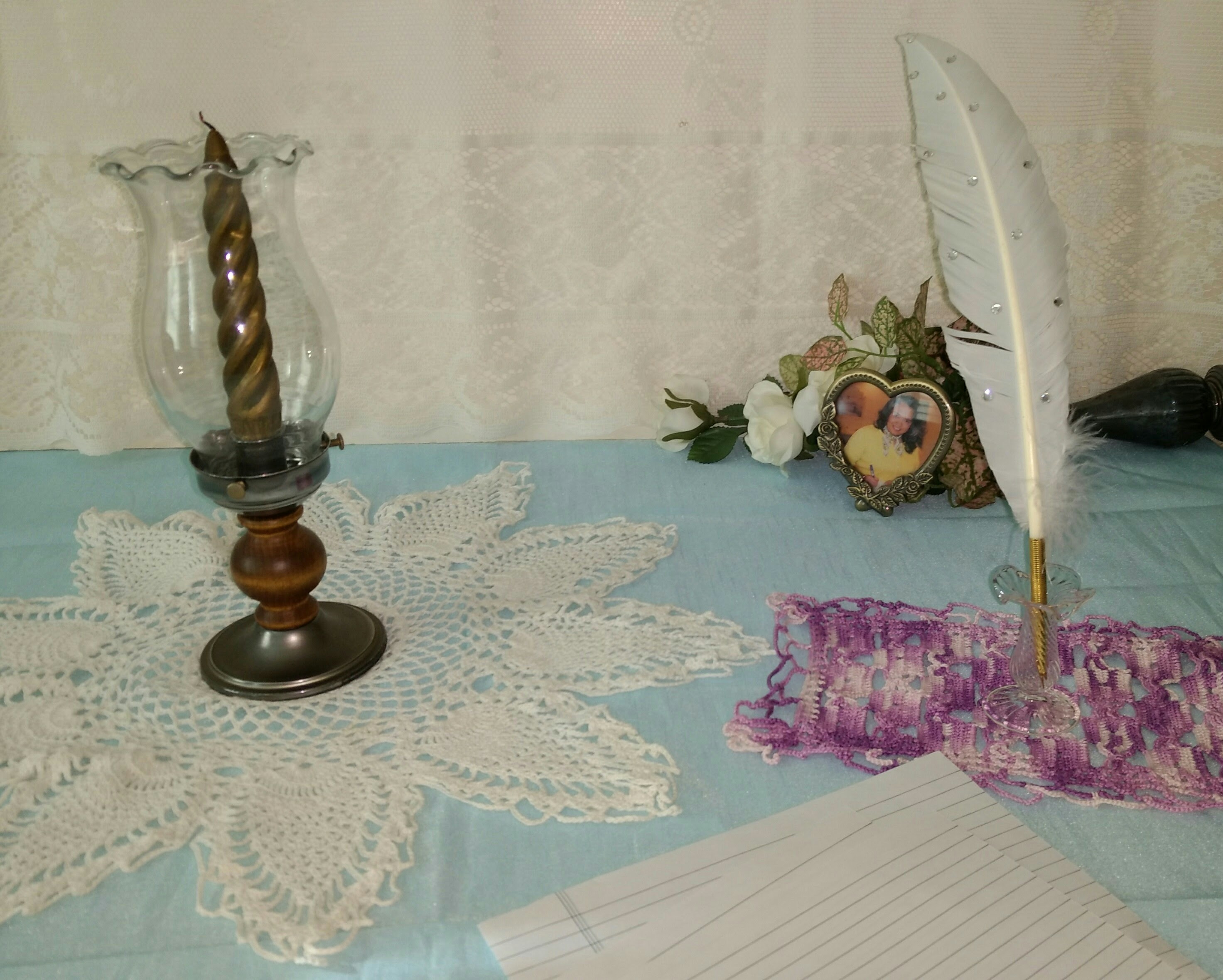 A still-life photograph featuring a lit brass candle holder, a white feather quill, a heart-shaped frame, and delicate lace atop a turquoise table.