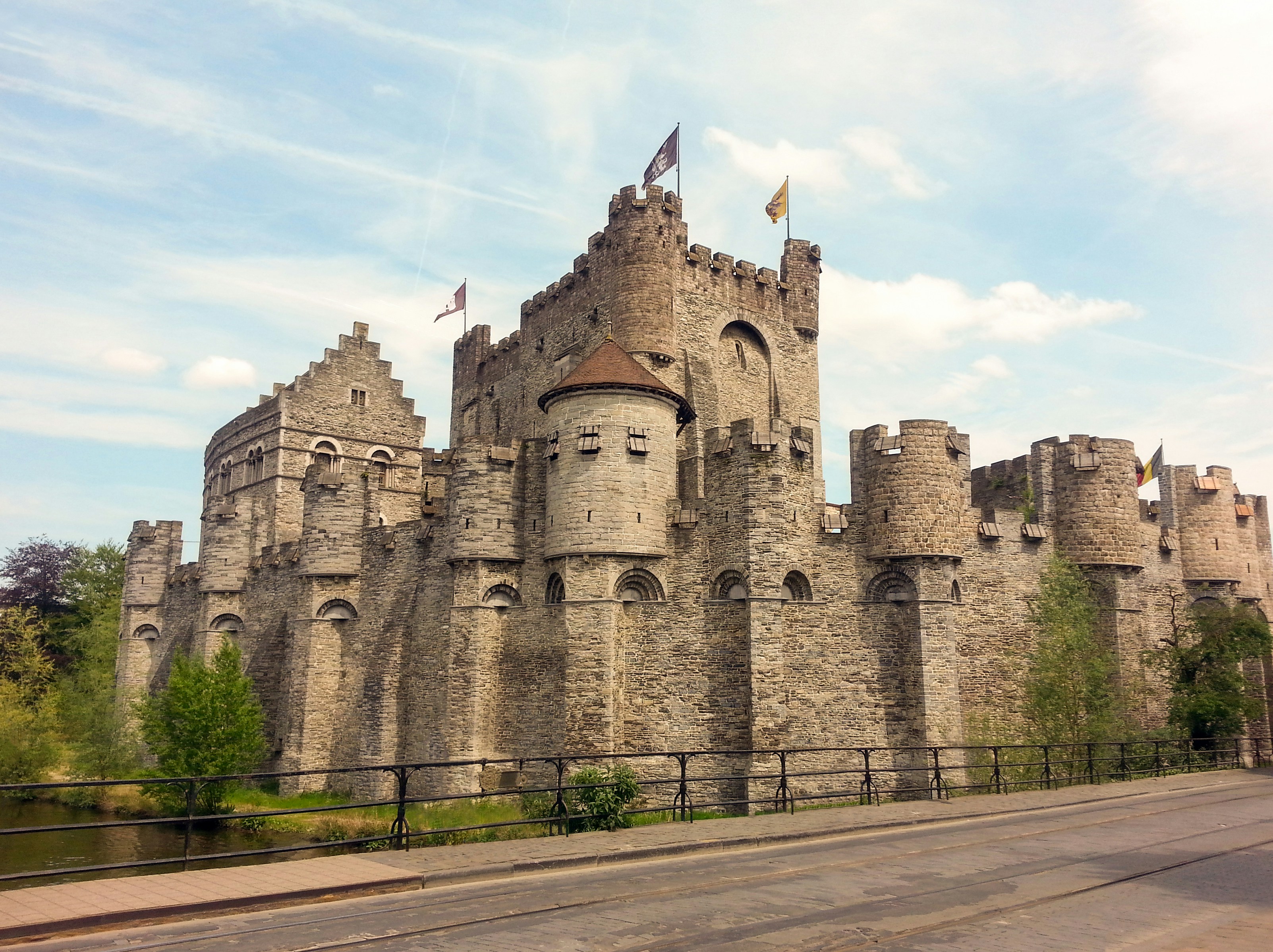 Sunlit medieval castle with crenellated towers and flags, set along a paved street with a railing and trees in the foreground.