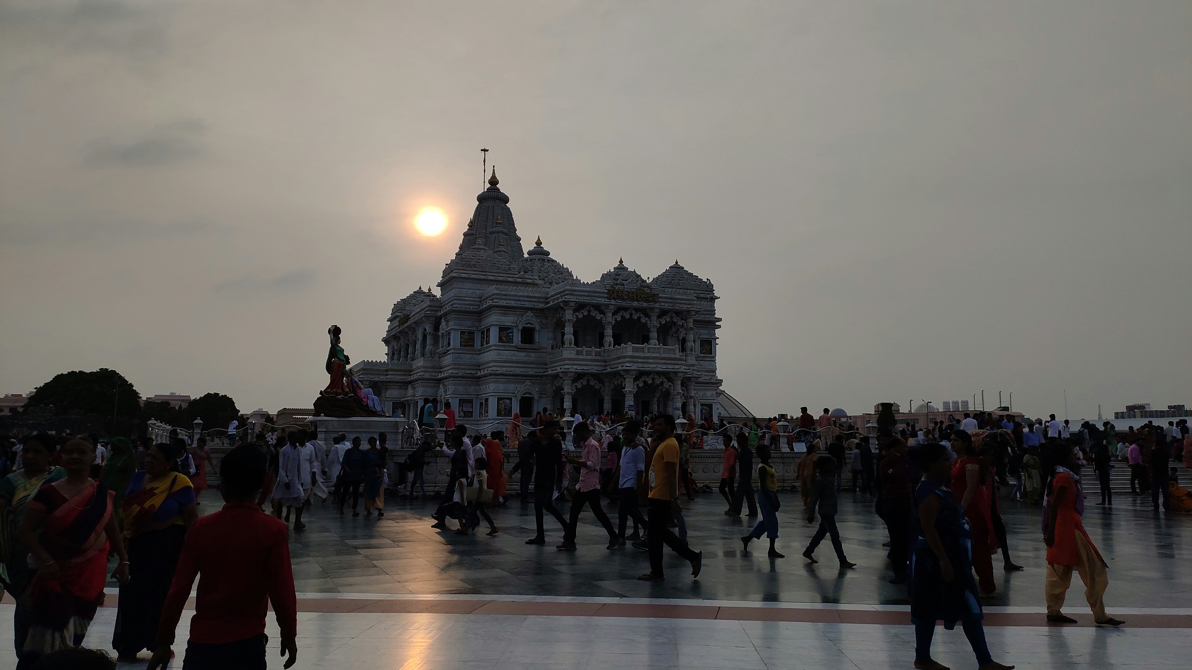 Crowd walking near a large temple with the sun setting behind overcast skies.